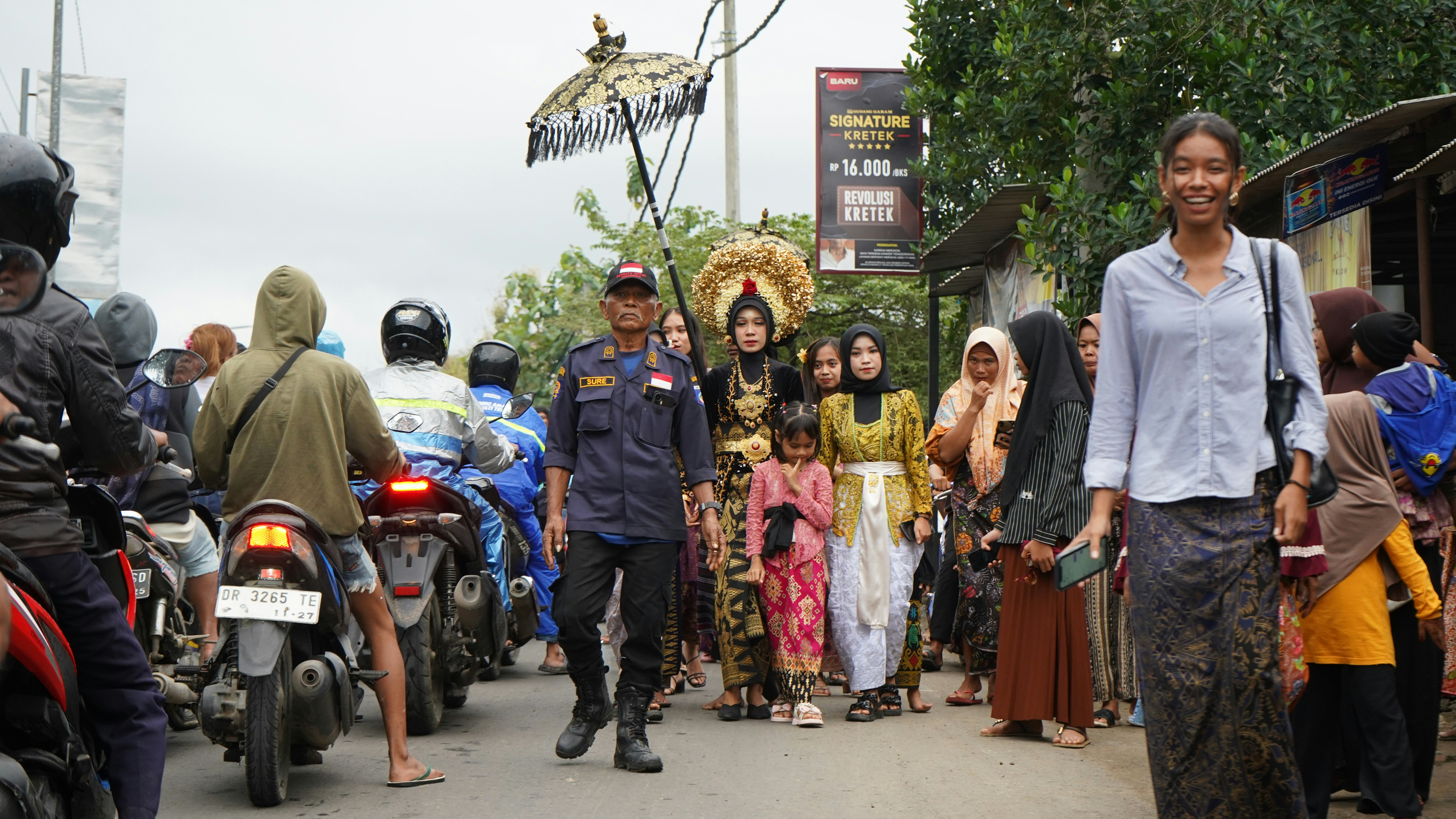 A Sasak bridal procession moves through village traffic, blending tradition and daily life in Lombok, Indonesia. A woman smiles at the camera in the foreground. | People walk in a parade on a crowded street.
