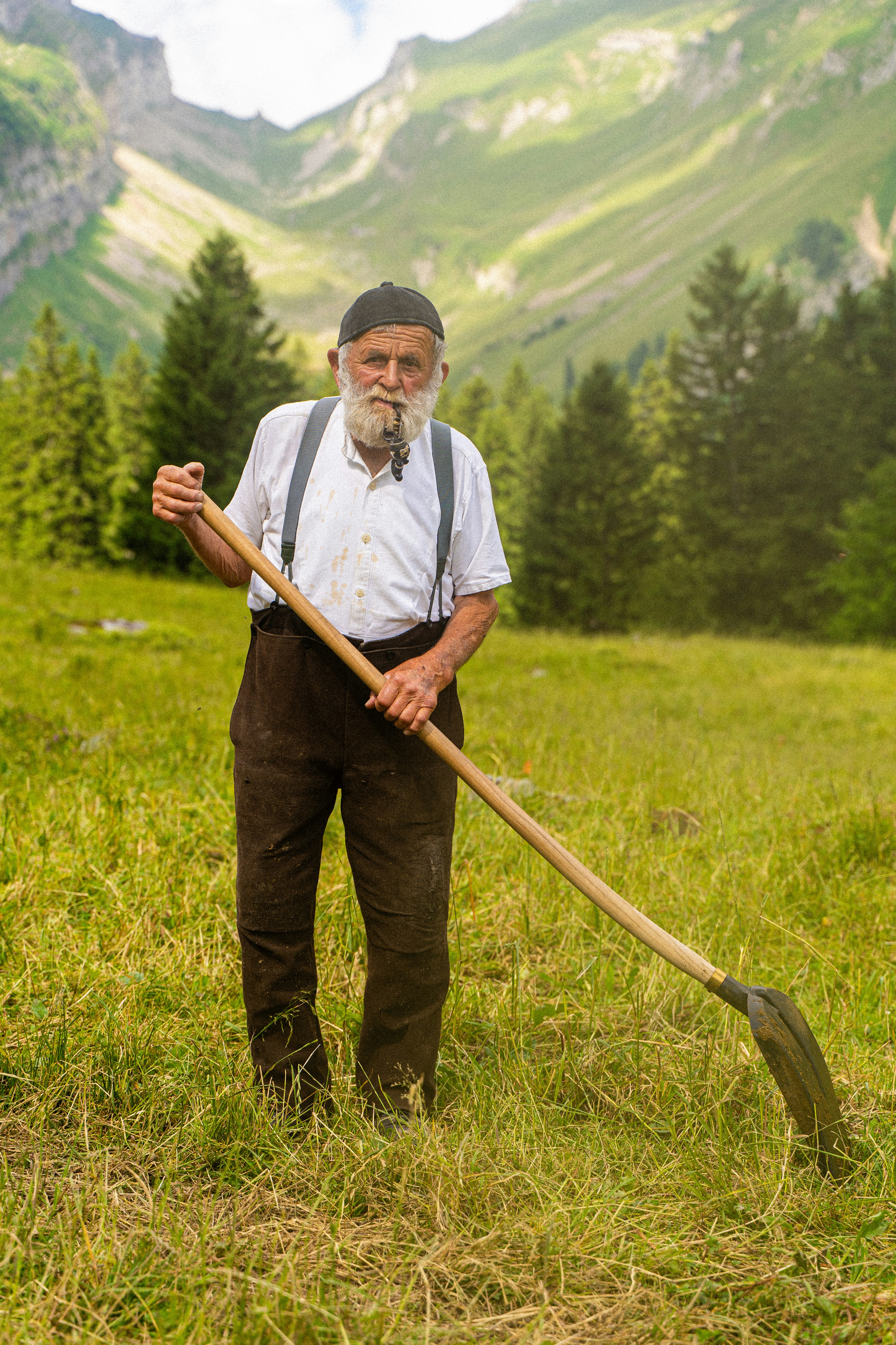 Elderly farmer stands with a scythe in a grassy field.