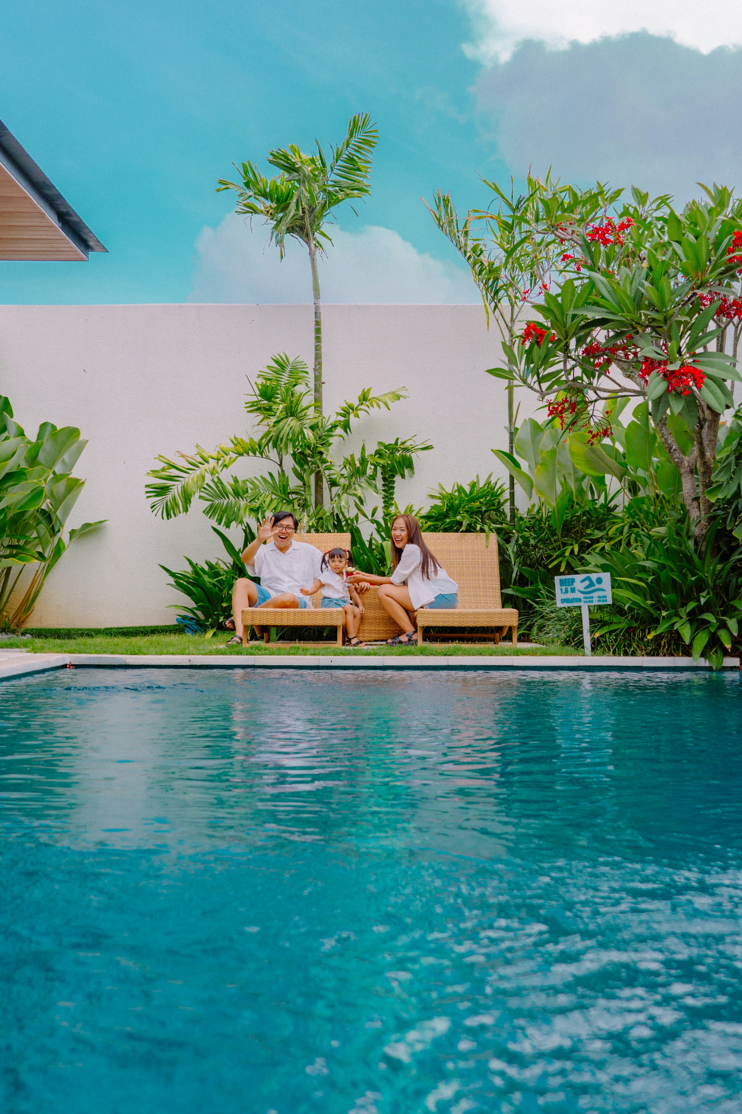 A happy family enjoys a relaxing moment by the private pool of a tropical villa in Bali. Surrounded by lush plants and blue skies, the scene captures a joyful summer getaway. | A family enjoys a relaxing day by the pool.