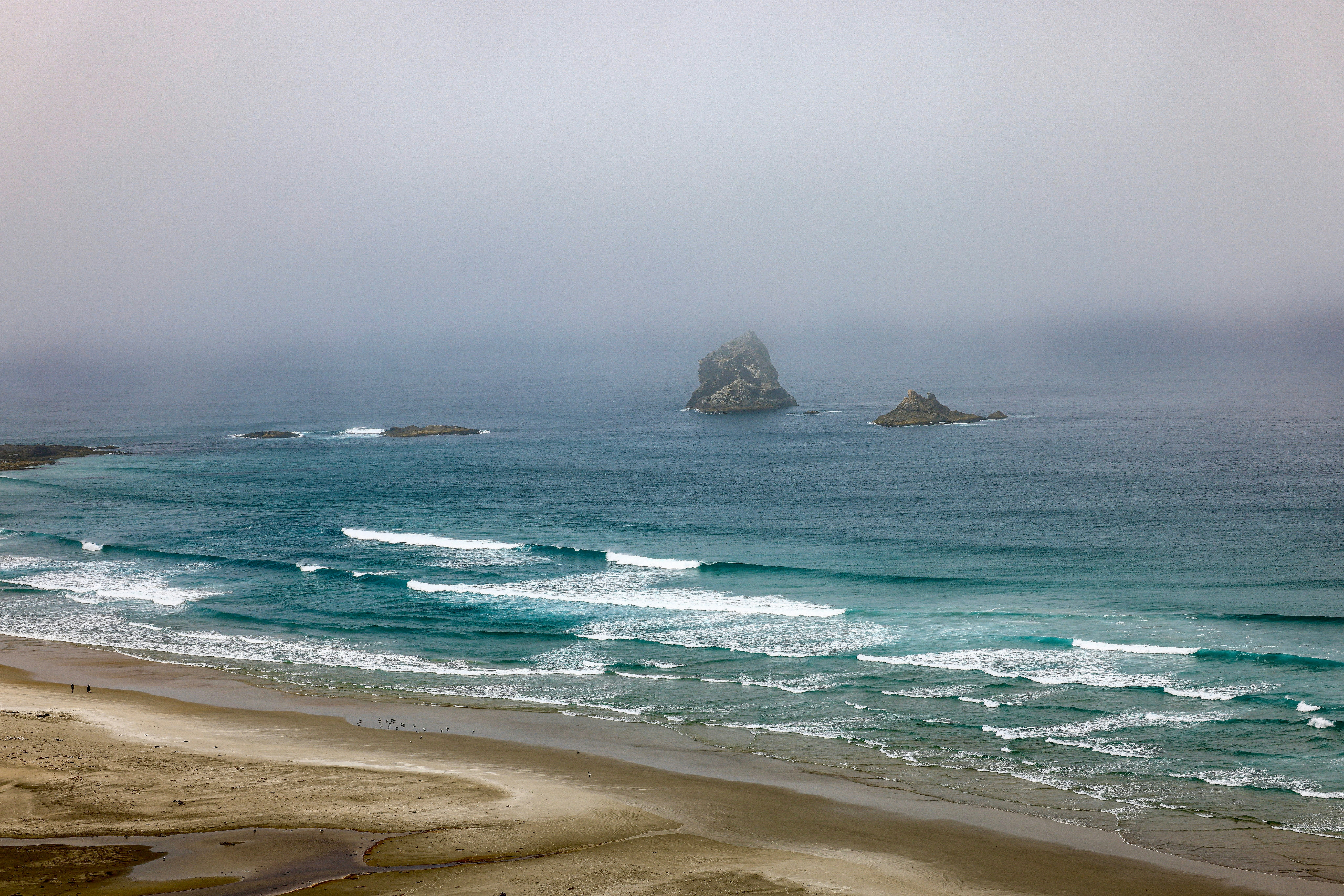 Misty coastal scene featuring gentle waves lapping against a sandy shore with distant rocky outcrops. The atmosphere evokes a sense of serenity and mystery.