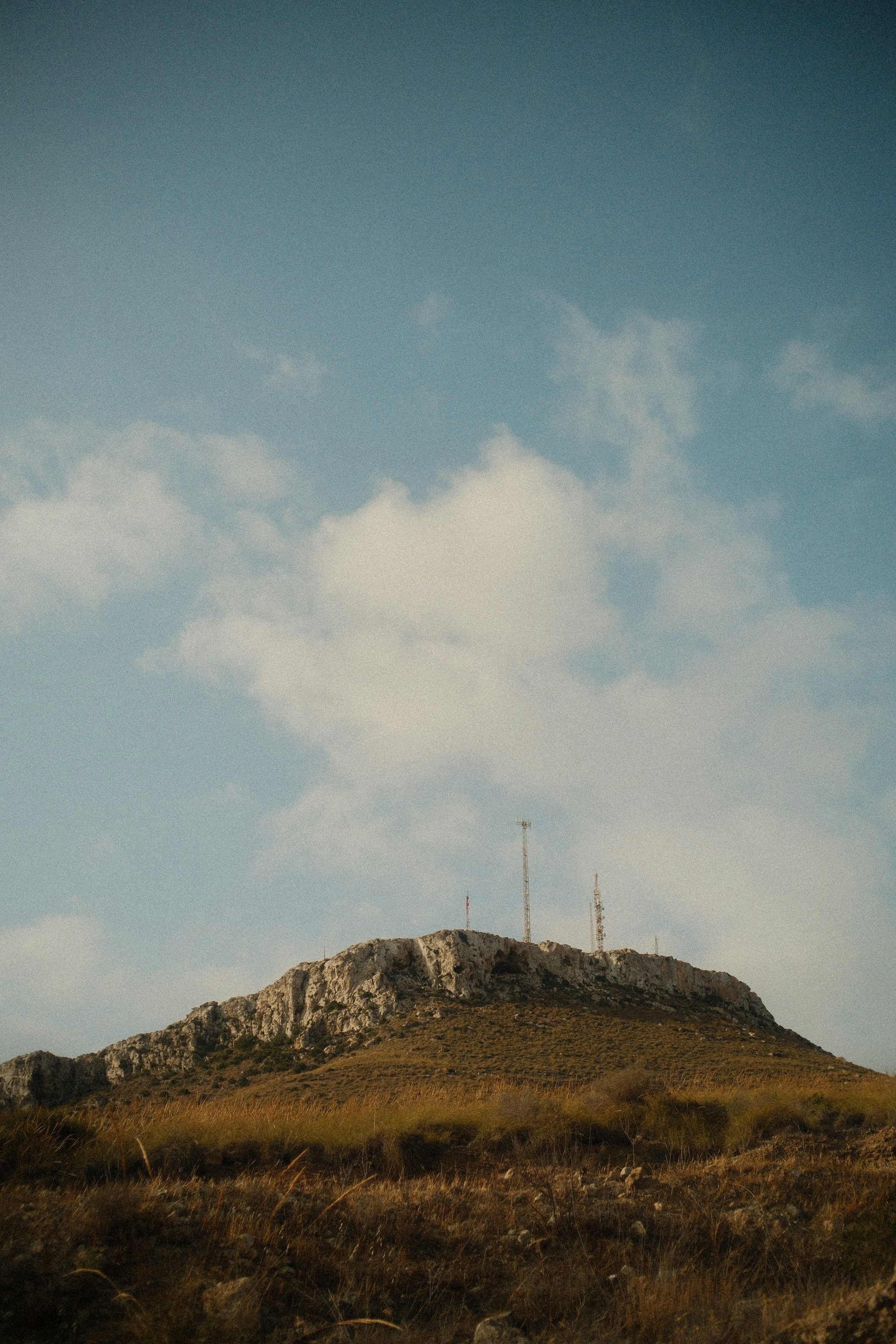 A warm, film-style photograph of a sunlit hill under a blue sky with scattered clouds. The hill's slope is covered in dry, golden-brown grass, leading up to a rocky summit where several communication antennas stand, blending nature with technology. The grainy texture gives the image a timeless, rustic feel. | A rocky hilltop stretches towards a blue sky.