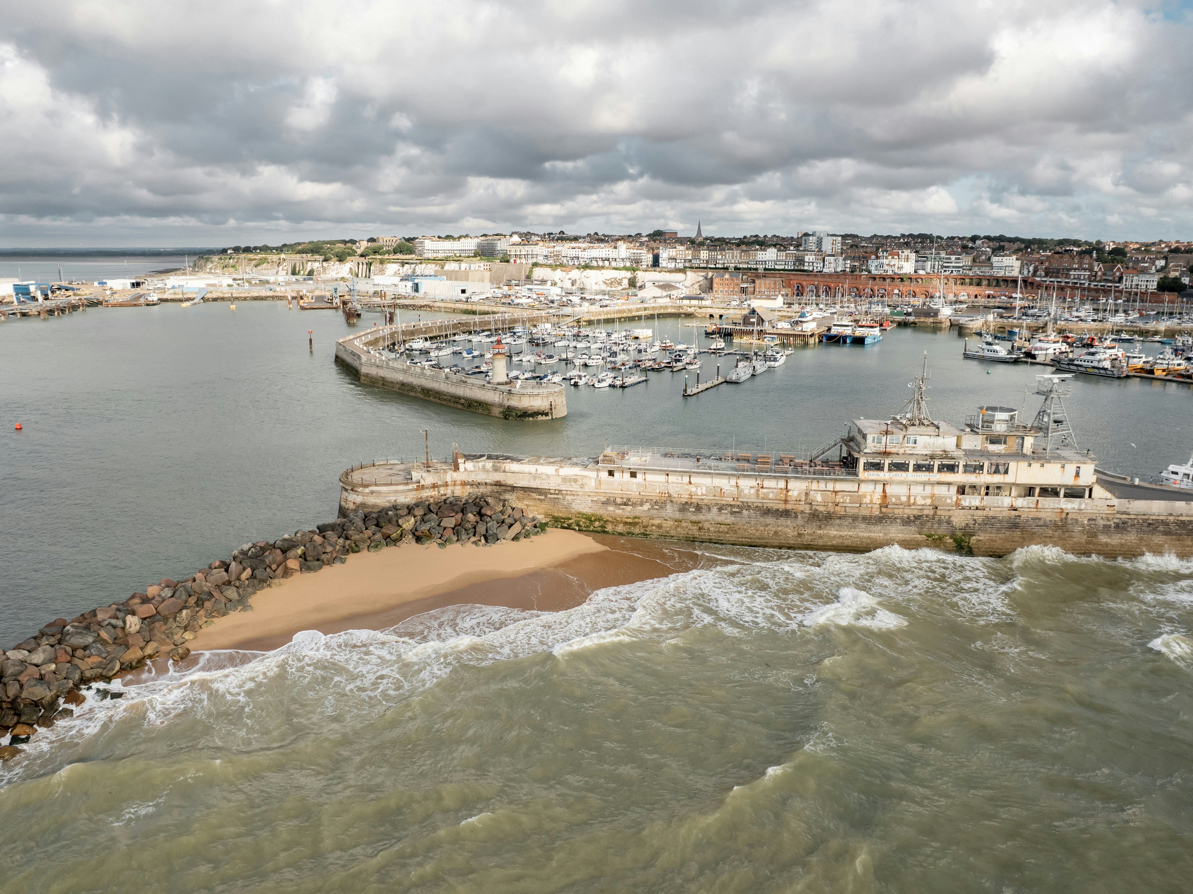 Aerial view of a bustling harbor with boats docked and waves gently lapping against the shore. The coastal town is framed by rolling clouds above.