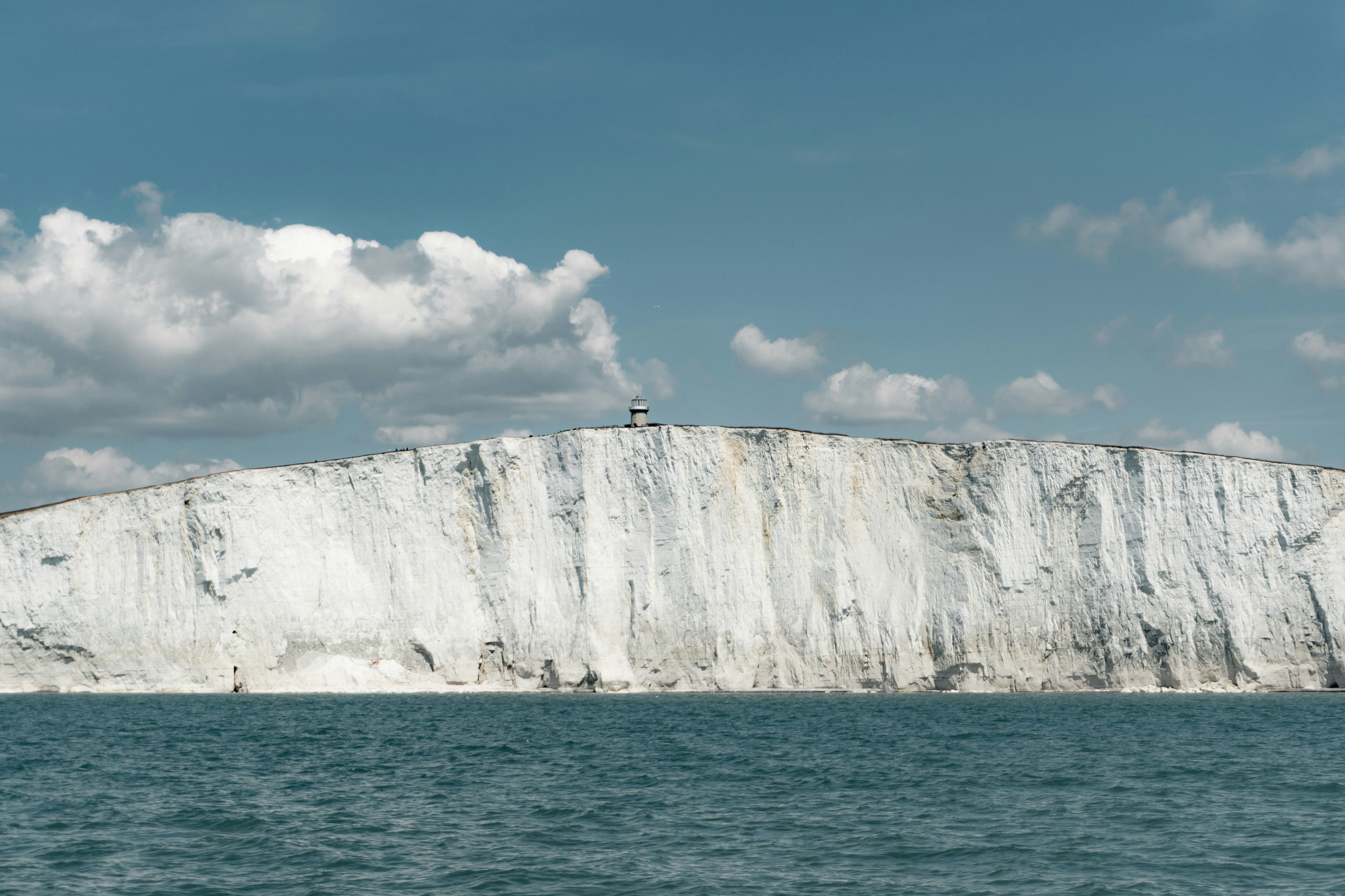 Majestic chalk cliffs rise sharply from the sea, with a lighthouse standing watch at the summit against a backdrop of fluffy clouds.