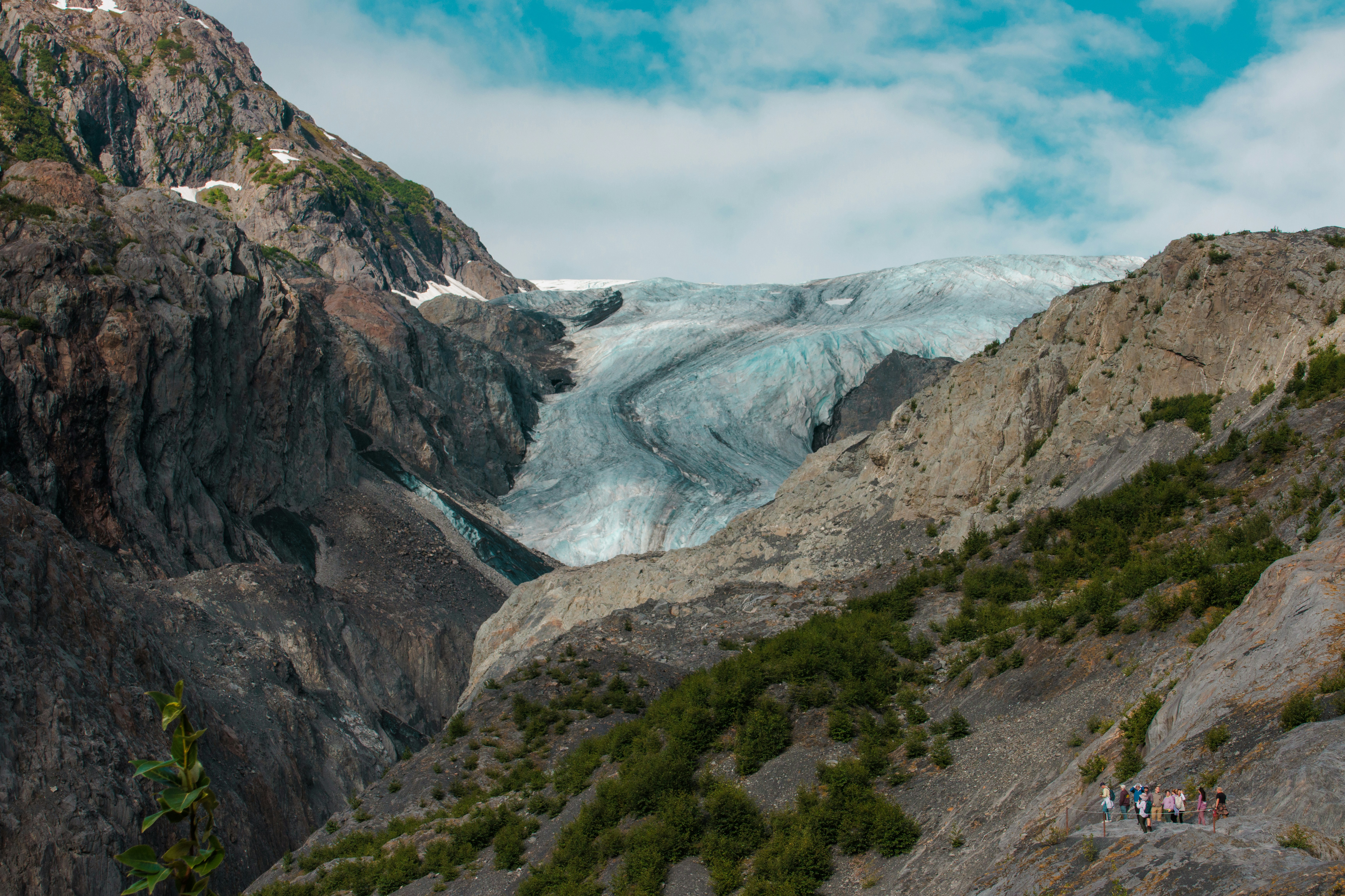 Expansive glacier flowing between rugged mountain peaks, with hikers visible on the rocky terrain below.