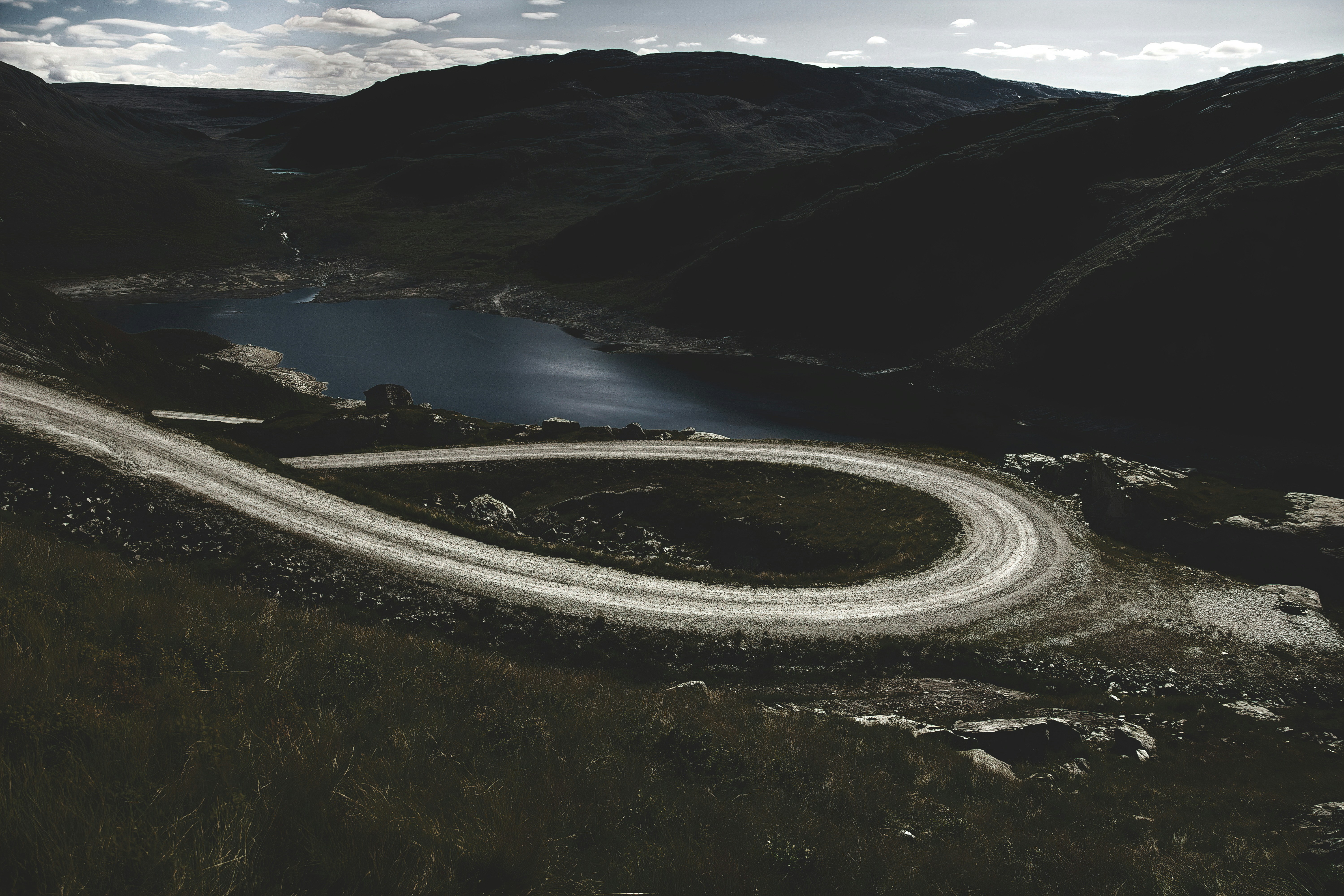 Curved dirt road meandering through rugged landscapes, leading to a serene body of water under a cloudy sky.