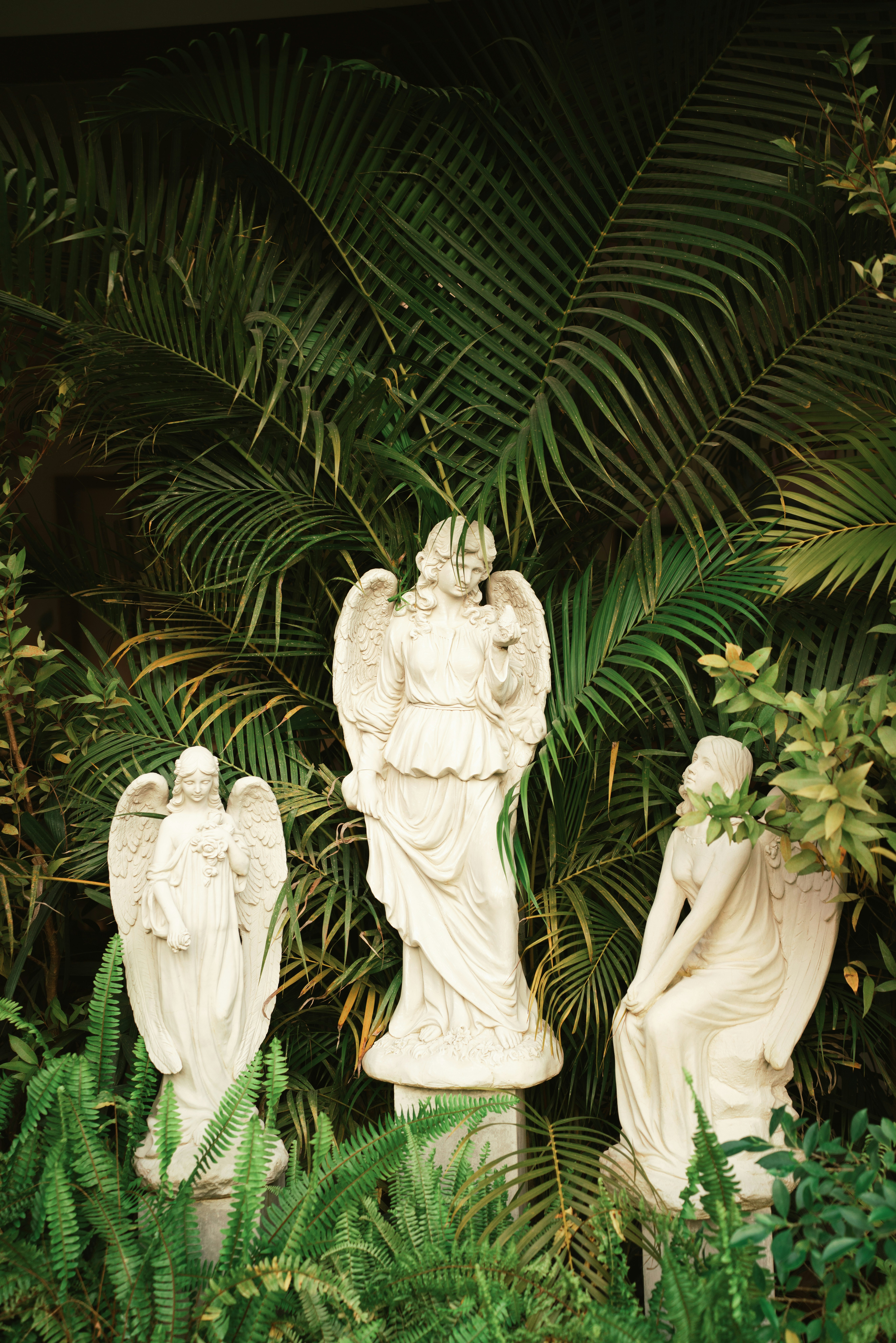 Three elegant white statues of angels nestled among lush green foliage, creating a serene atmosphere in a garden setting.