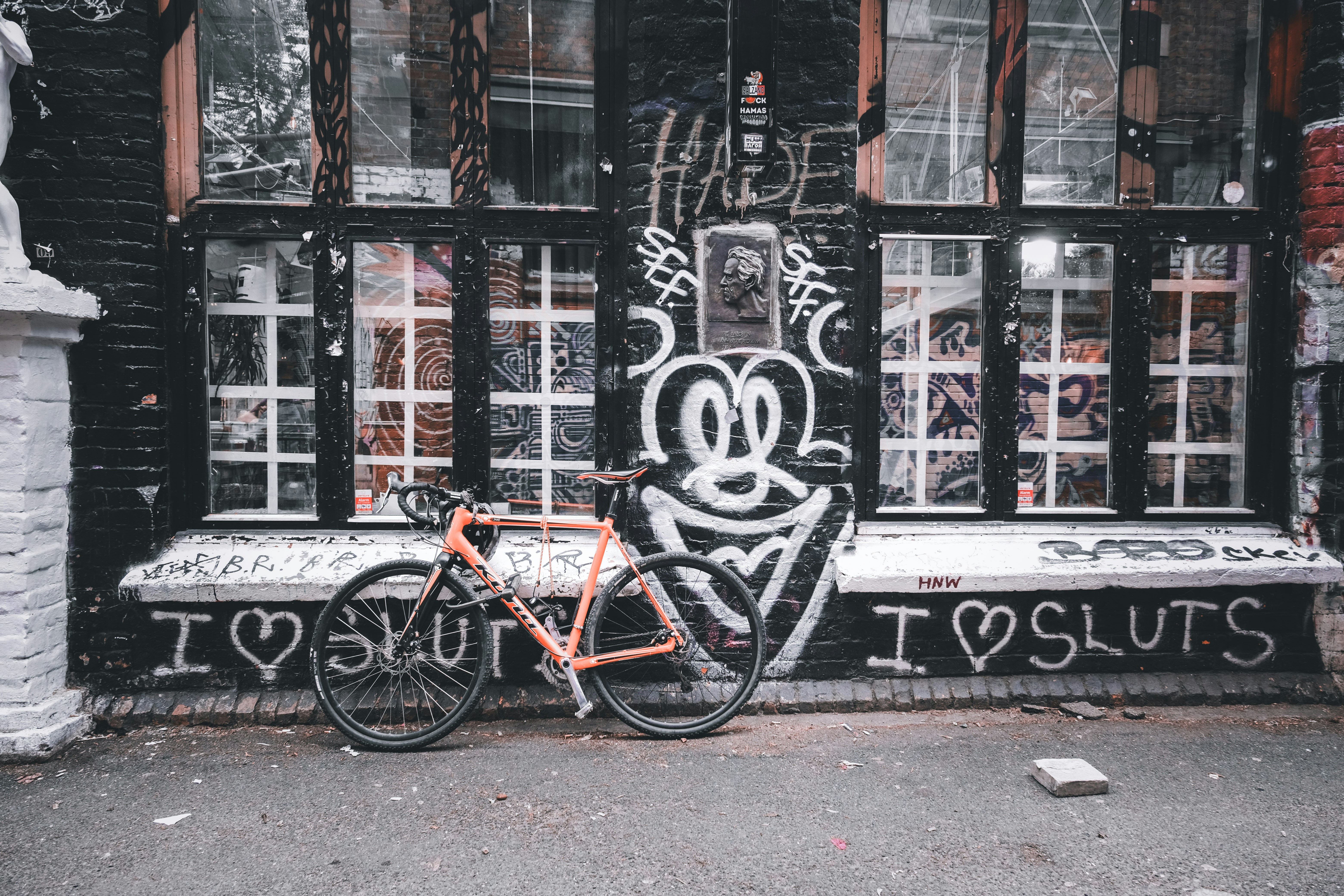 An orange bicycle rests against a graffiti-covered wall, featuring playful street art and vibrant messages. The scene captures the essence of urban culture and creativity.