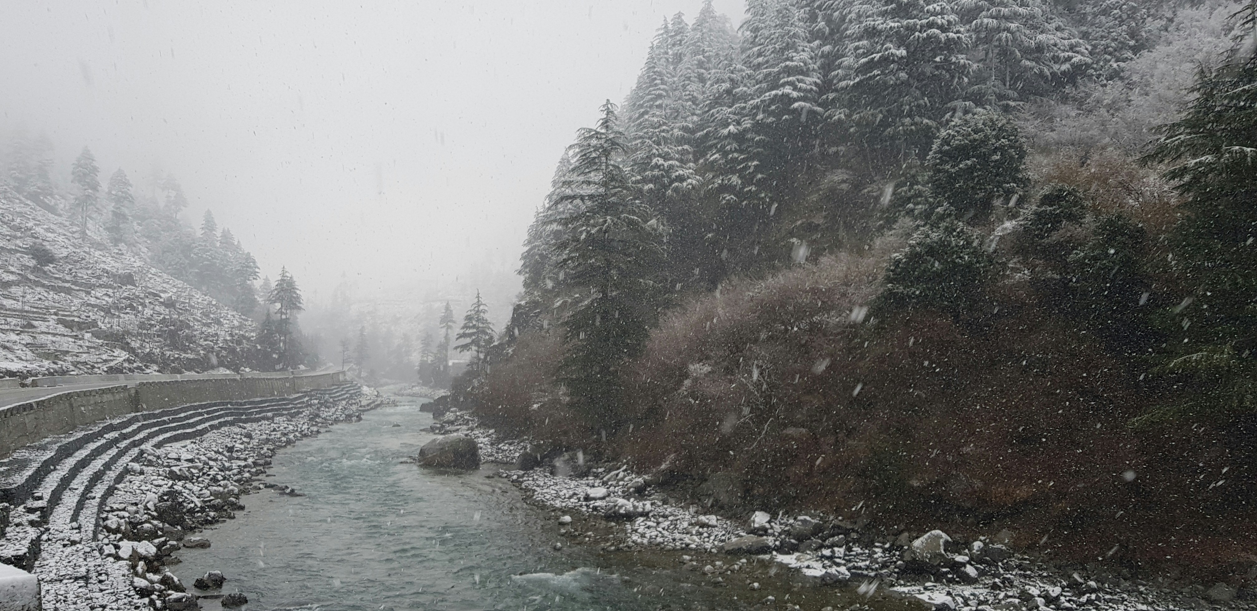 Snow falls over a river and snowy forest.