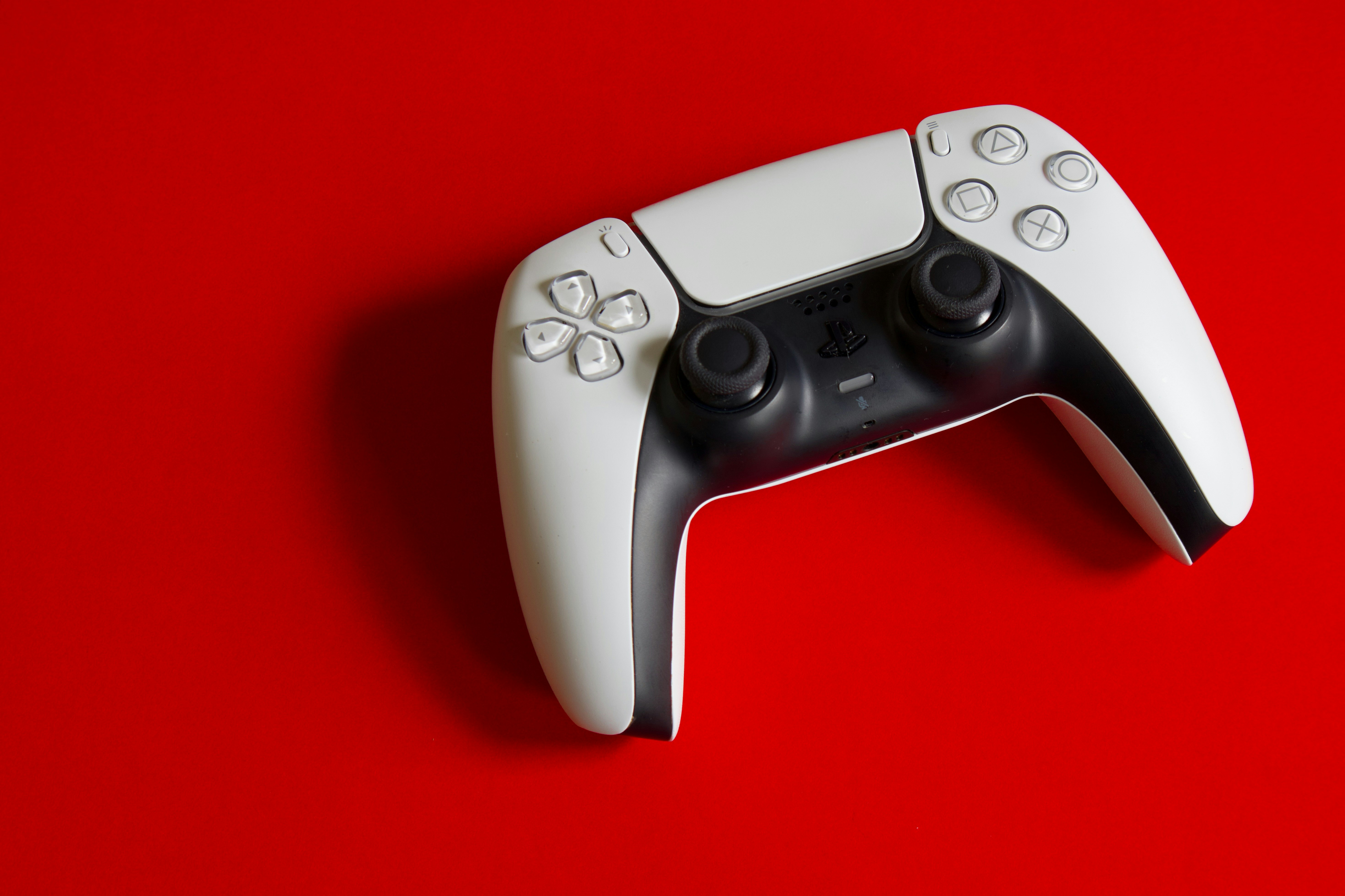 This striking image showcases a PlayStation 5 DualSense controller elegantly positioned against a bold red background. The sleek curves and modern design of the controller are highlighted by strong lighting, making it the central focus of the composition. The vivid color contrast and minimalistic setting evoke a sense of excitement and innovation, ideal for conveying themes of advanced gaming and technology. Perfect for use in presentations, articles, advertisements, or digital content centered on gaming, technology trends, or product reviews. | Playstation 5 dualsense controller on a red surface.