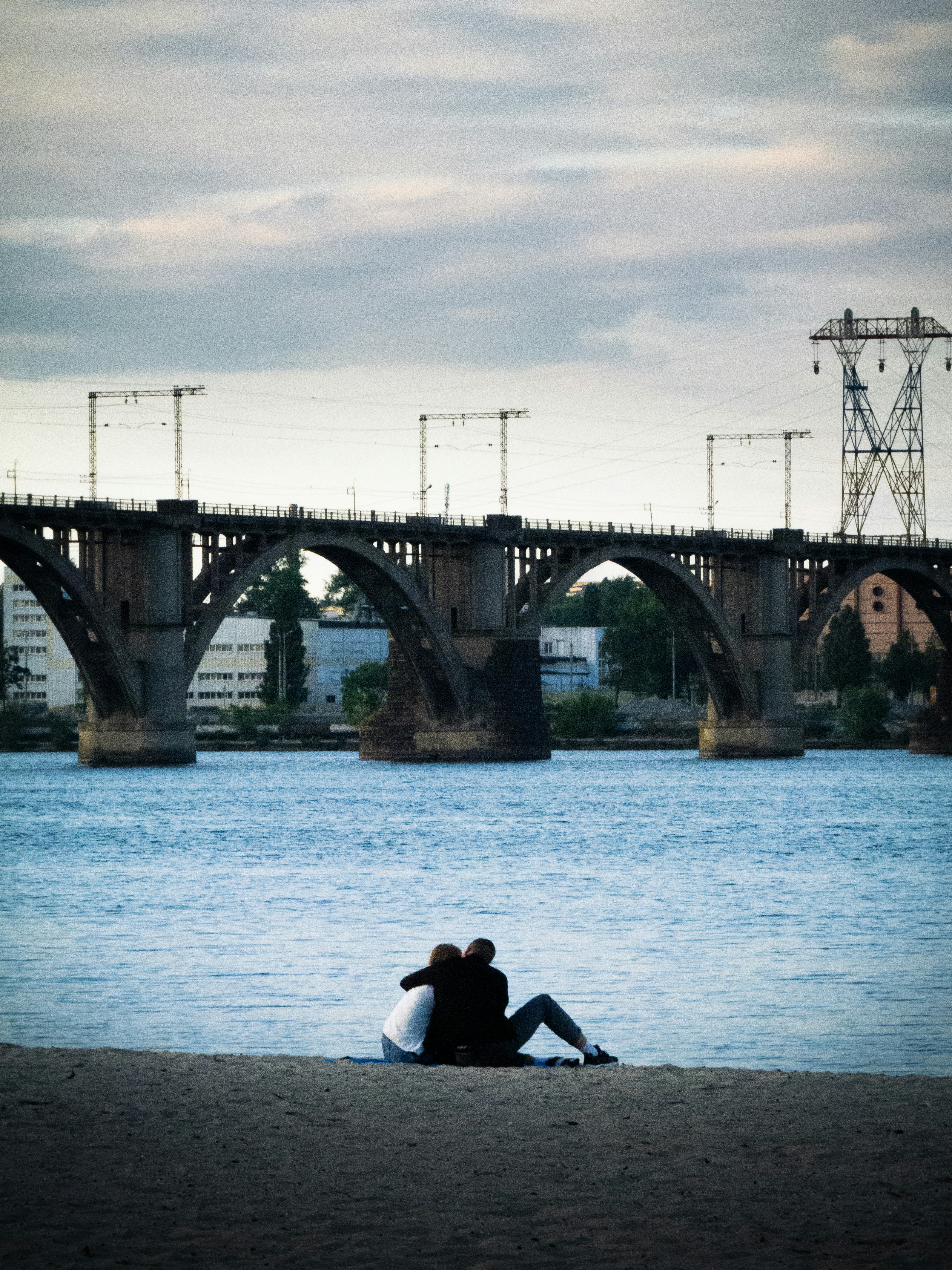 couple cuddling and a bridge. | Couple sits by the water, bridge in the background.