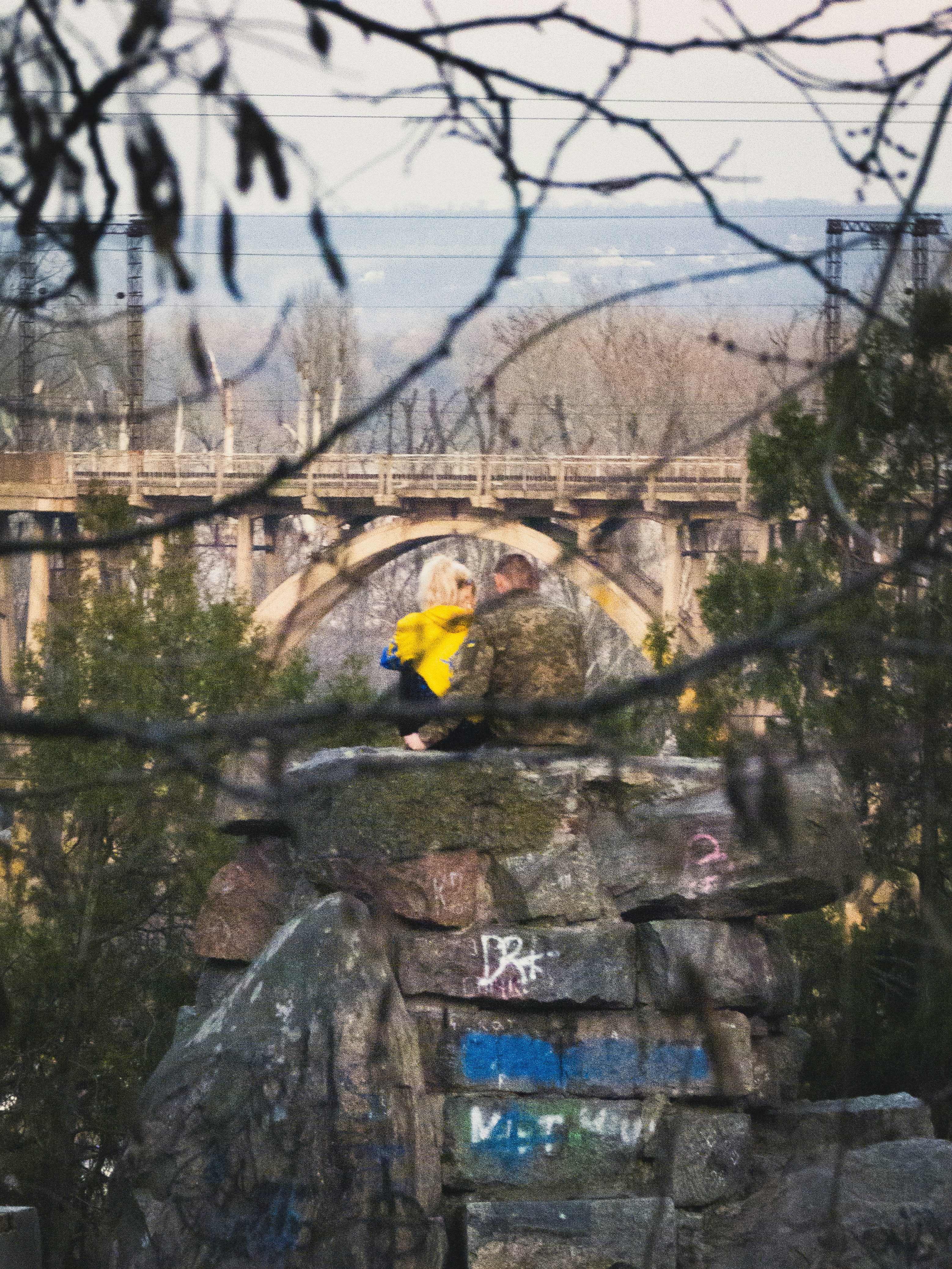 Couple seated on a rocky outcrop, surrounded by trees and distant structures, sharing a moment of intimacy. Graffiti adorns the rocks, adding character to the scene.