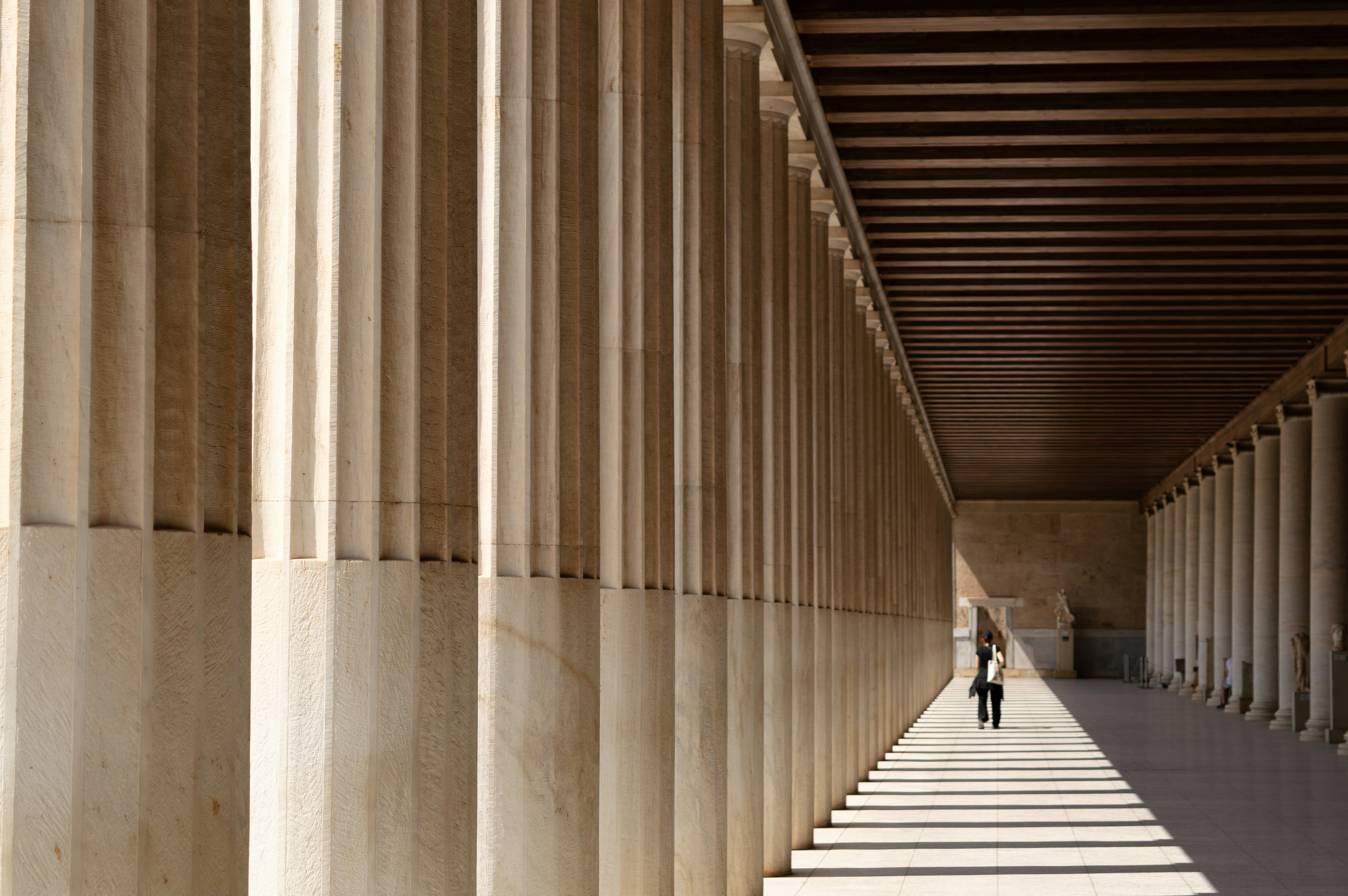 A solitary figure walks through a corridor lined with towering columns, showcasing the grandeur of classical design. The interplay of light and shadow enhances the structural elegance.