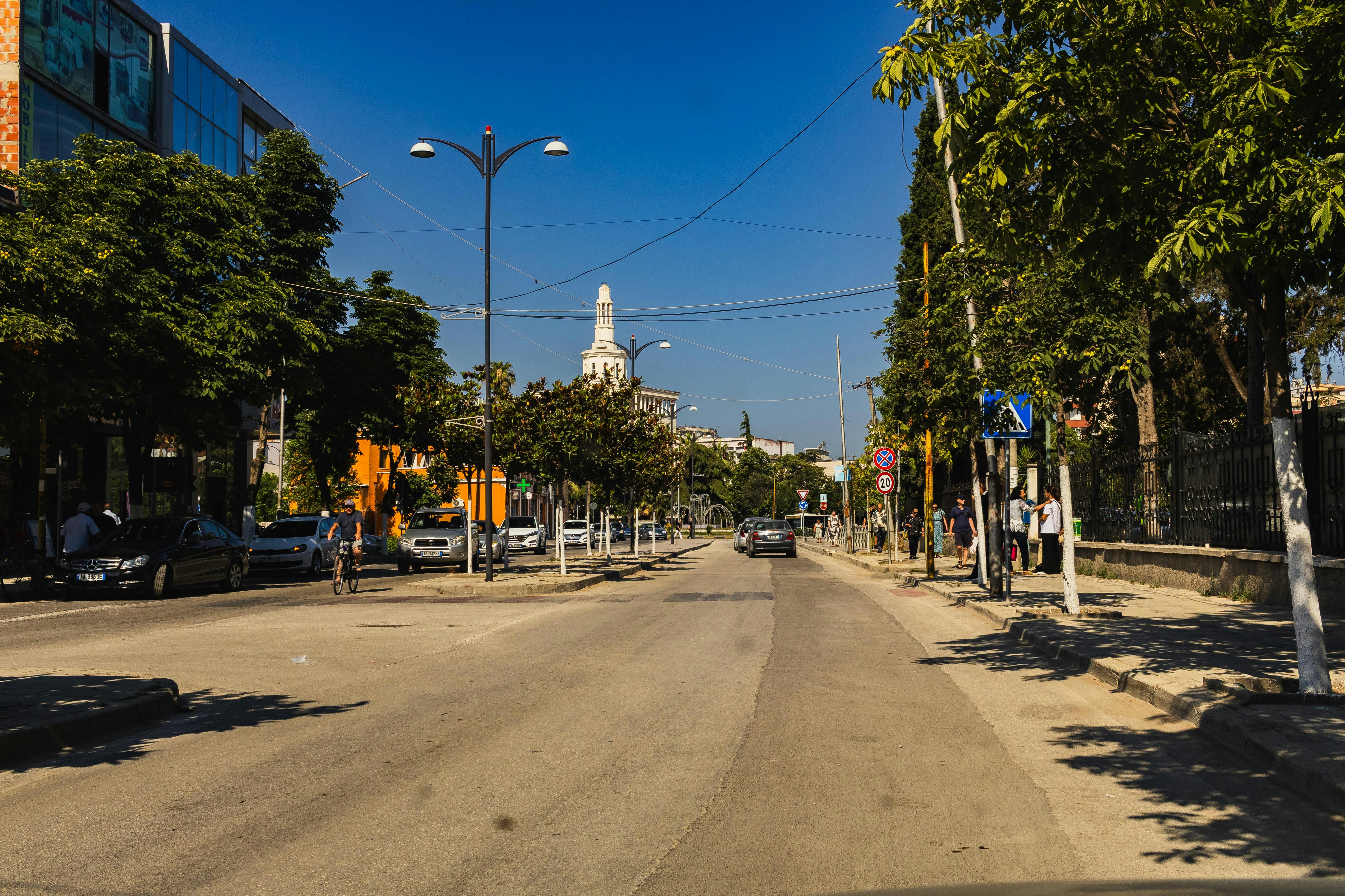 A sunlit street scene showcasing a blend of modern architecture and vibrant greenery, with vehicles and pedestrians navigating the urban landscape.