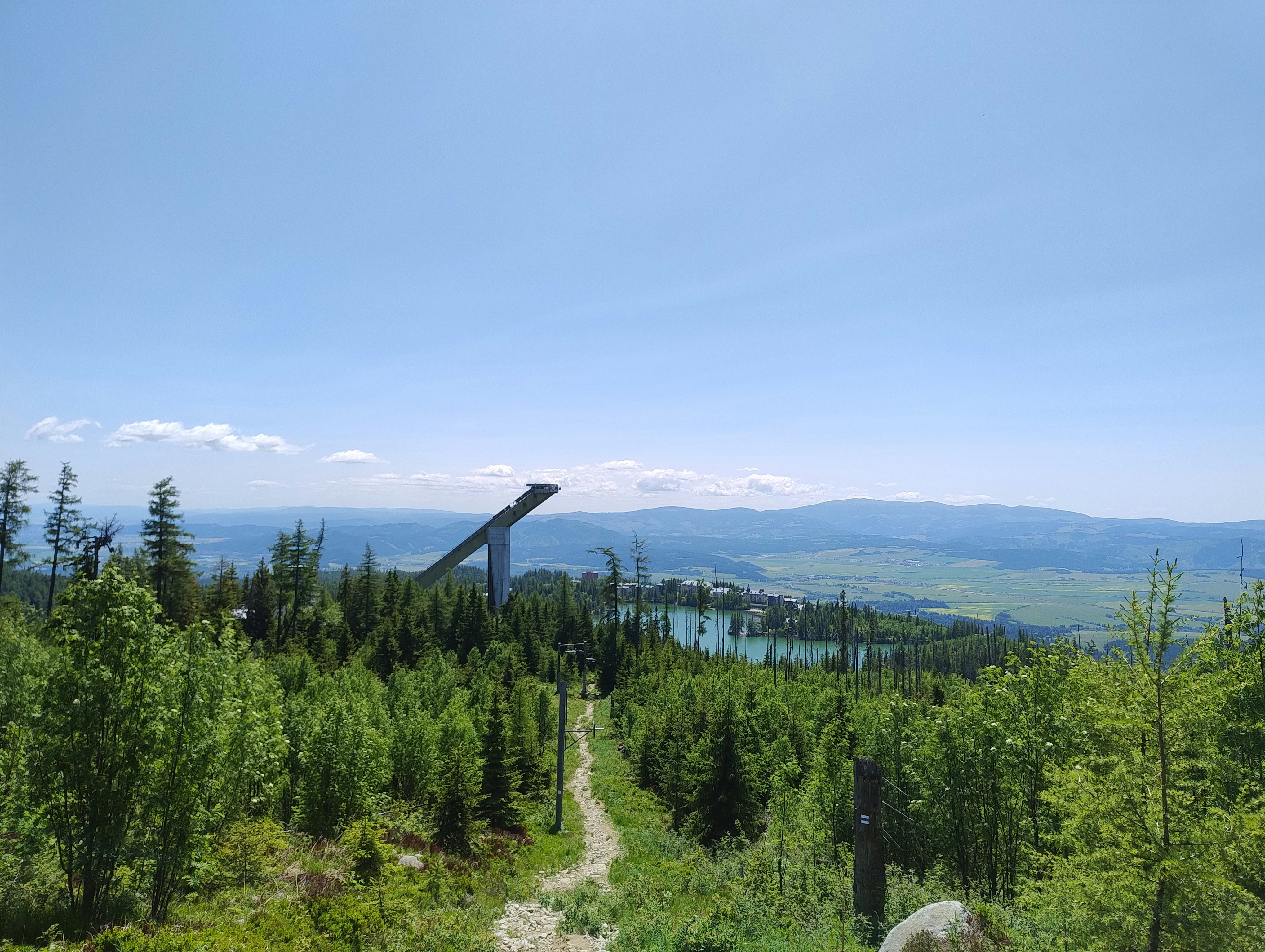 Ski jump structure overlooking a lush forested landscape and serene lake, framed by distant mountains under a clear blue sky.