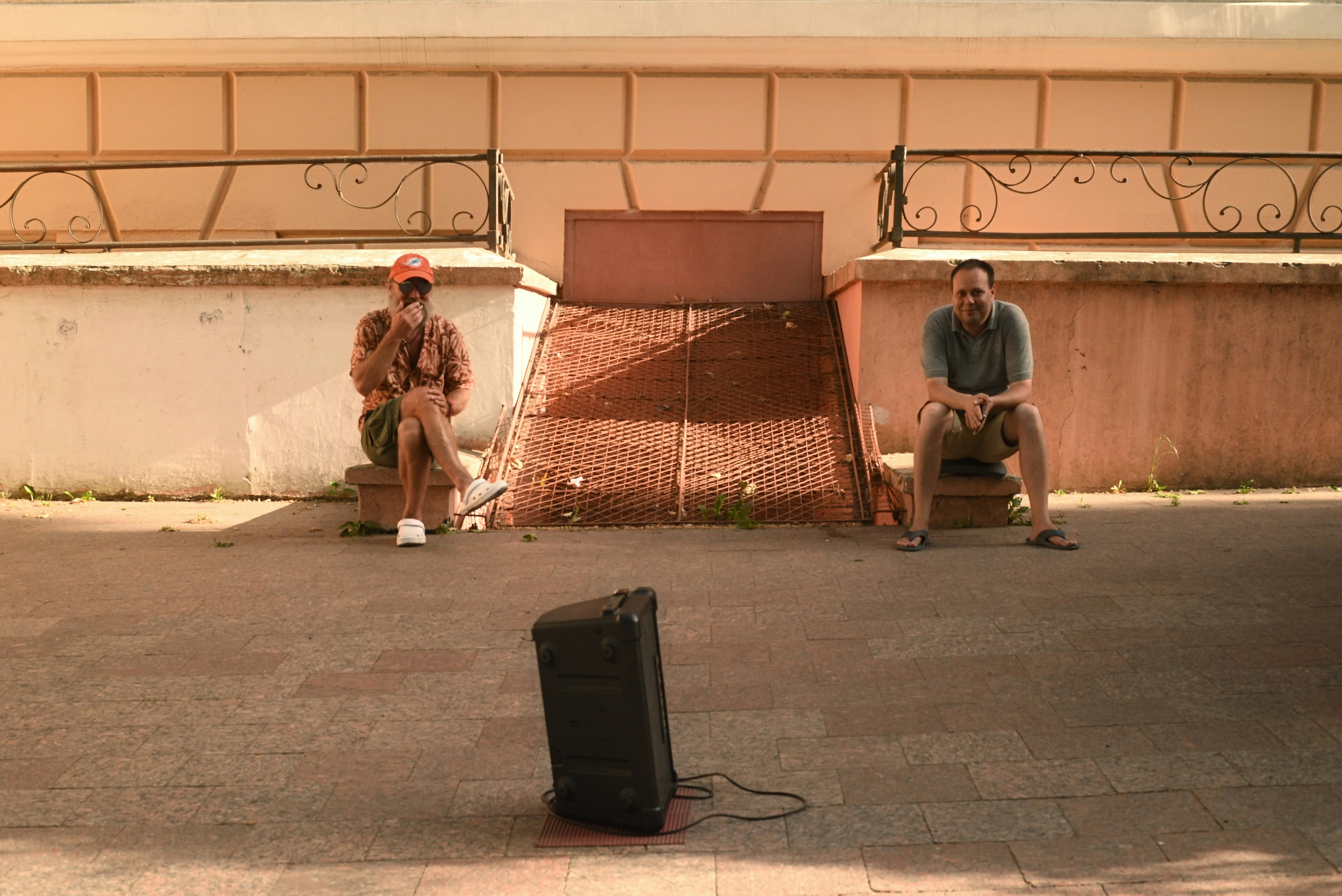 Two men sit apart on opposite sides of a sloped, gridded metal structure, possibly an entrance to an underground area, against a light-colored building. In the foreground, a single black speaker rests on the paved ground between them, suggesting a musical performance or gathering. The scene has a casual, outdoor urban feel, possibly on a sunny day given the lighting. | Two people sit with a speaker between them.