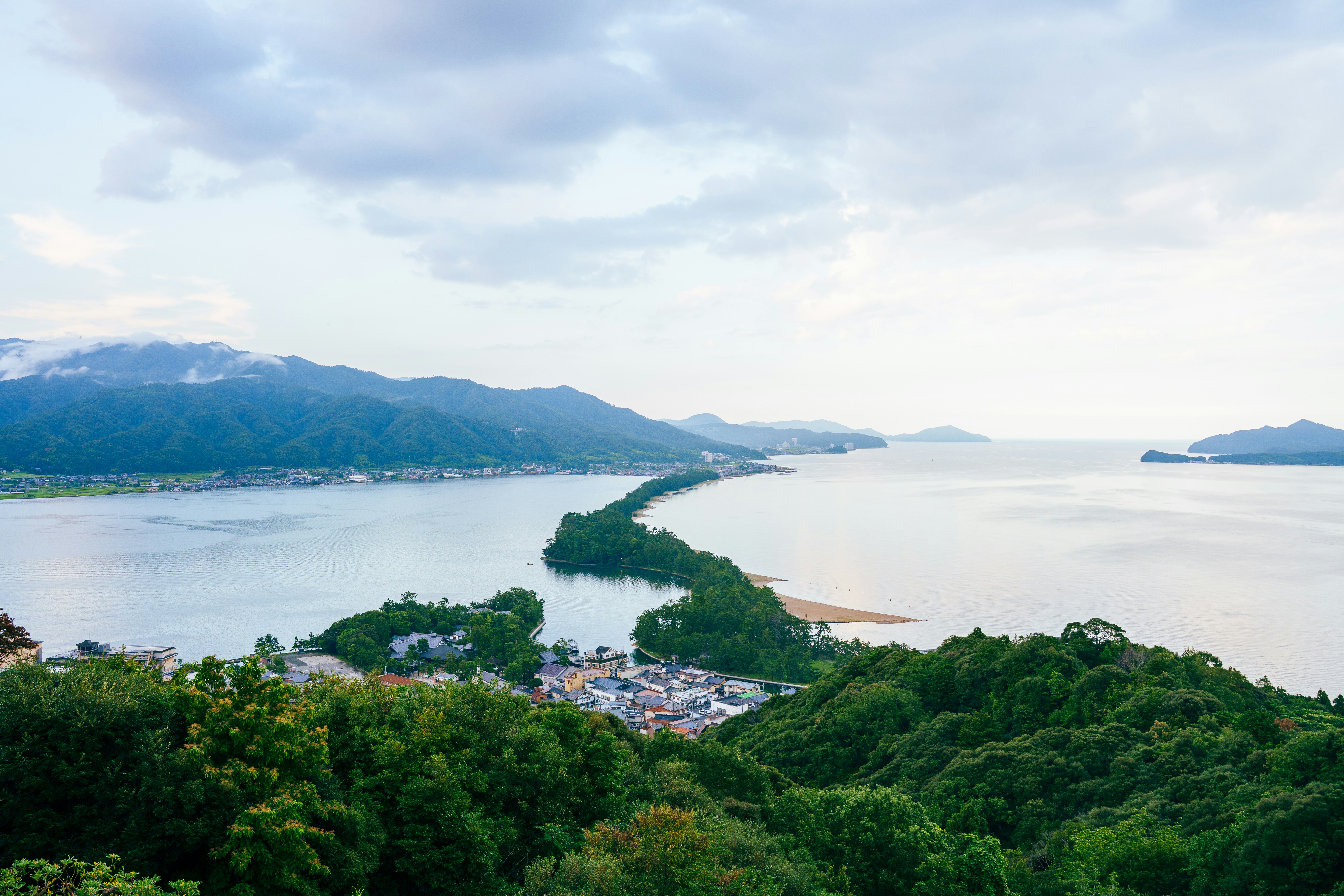 A panoramic view of a coastal inlet surrounded by lush greenery and distant mountains, showcasing the tranquil waters and the meandering landforms.