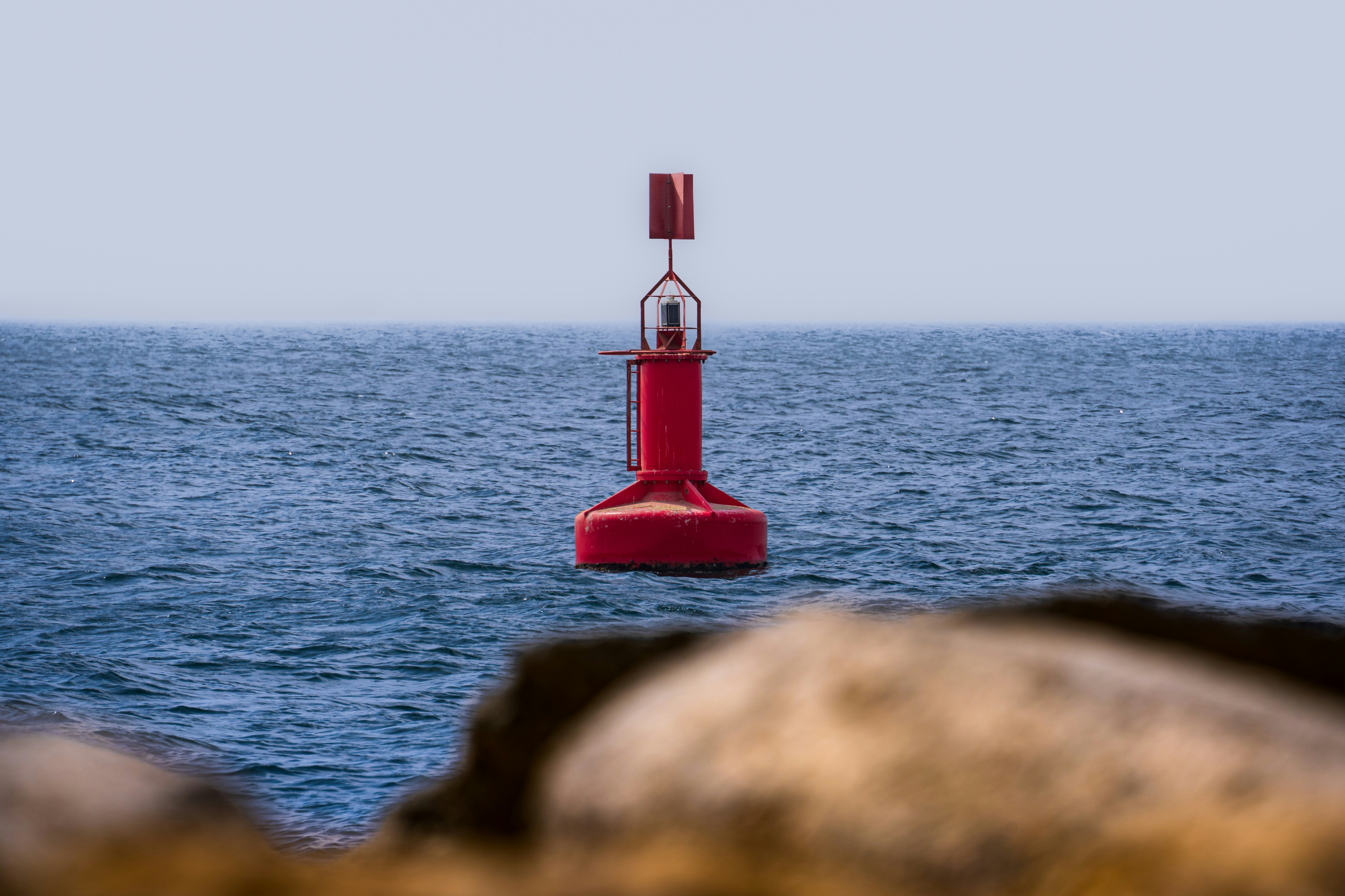A solitary red buoy stands prominently in the ocean, surrounded by gentle waves under a clear sky. The foreground features blurred rocks, adding depth to the scene.
