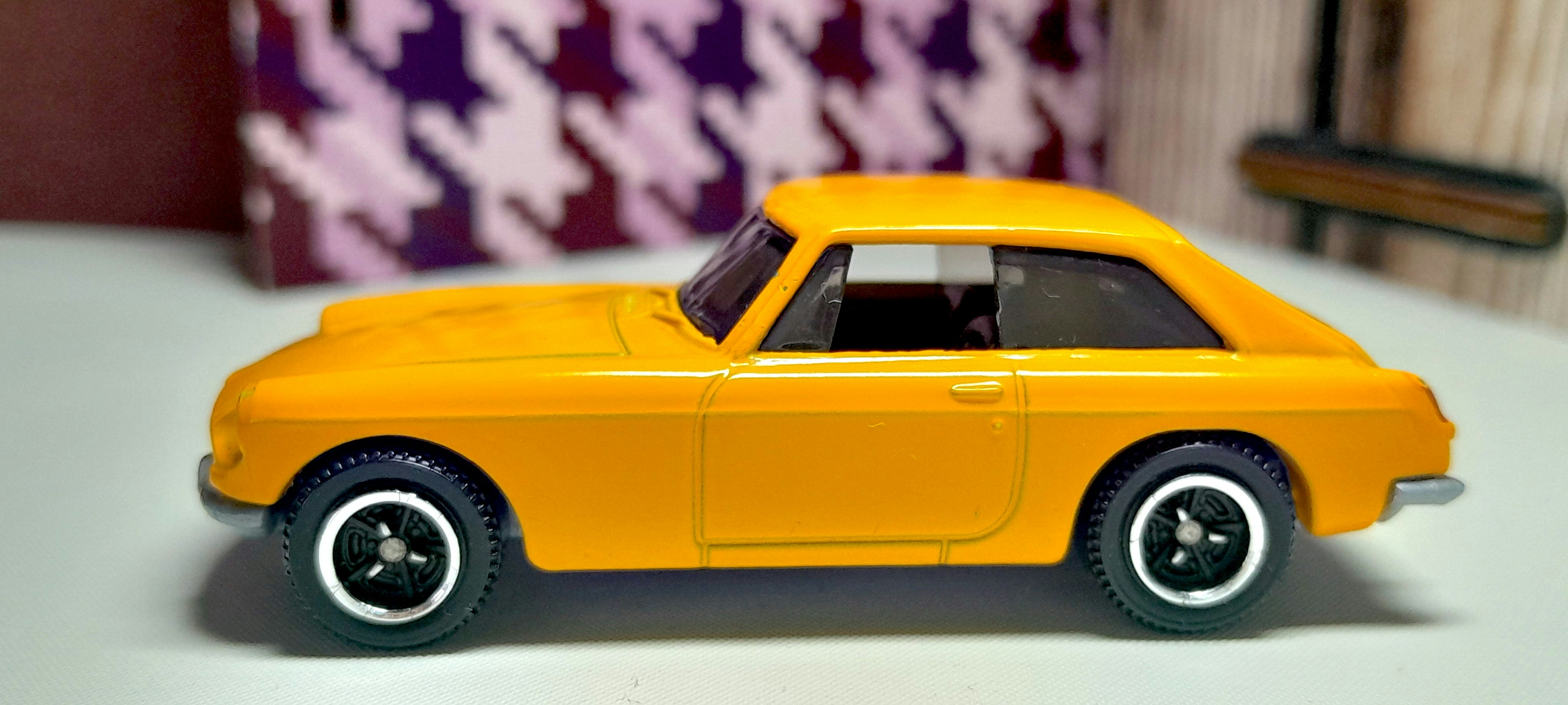 A bright yellow toy car model, showcasing classic design elements, positioned on a neutral surface with a patterned backdrop.