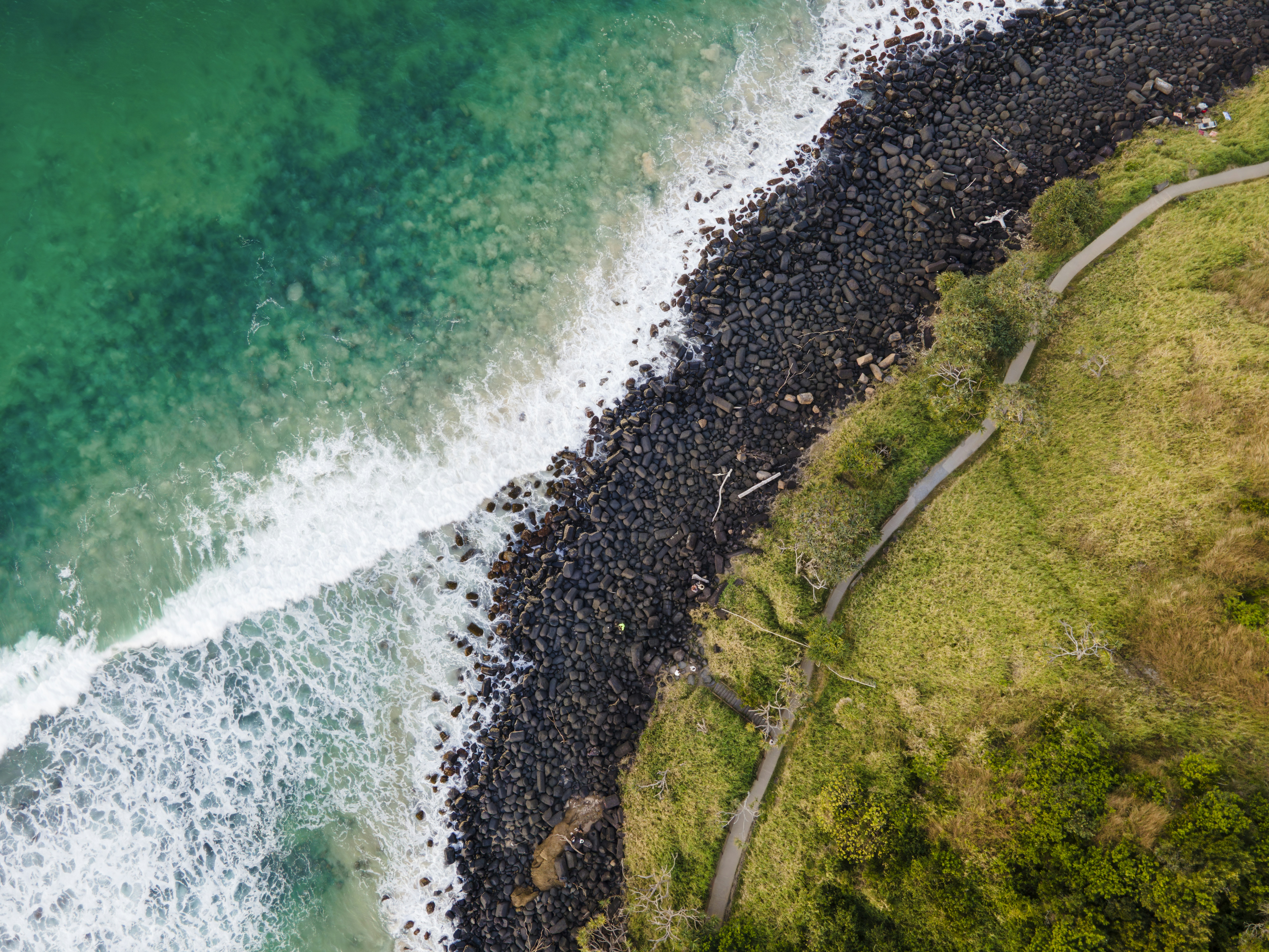 Aerial view of rocky shoreline meeting vibrant turquoise waters, with a winding path leading through lush greenery.