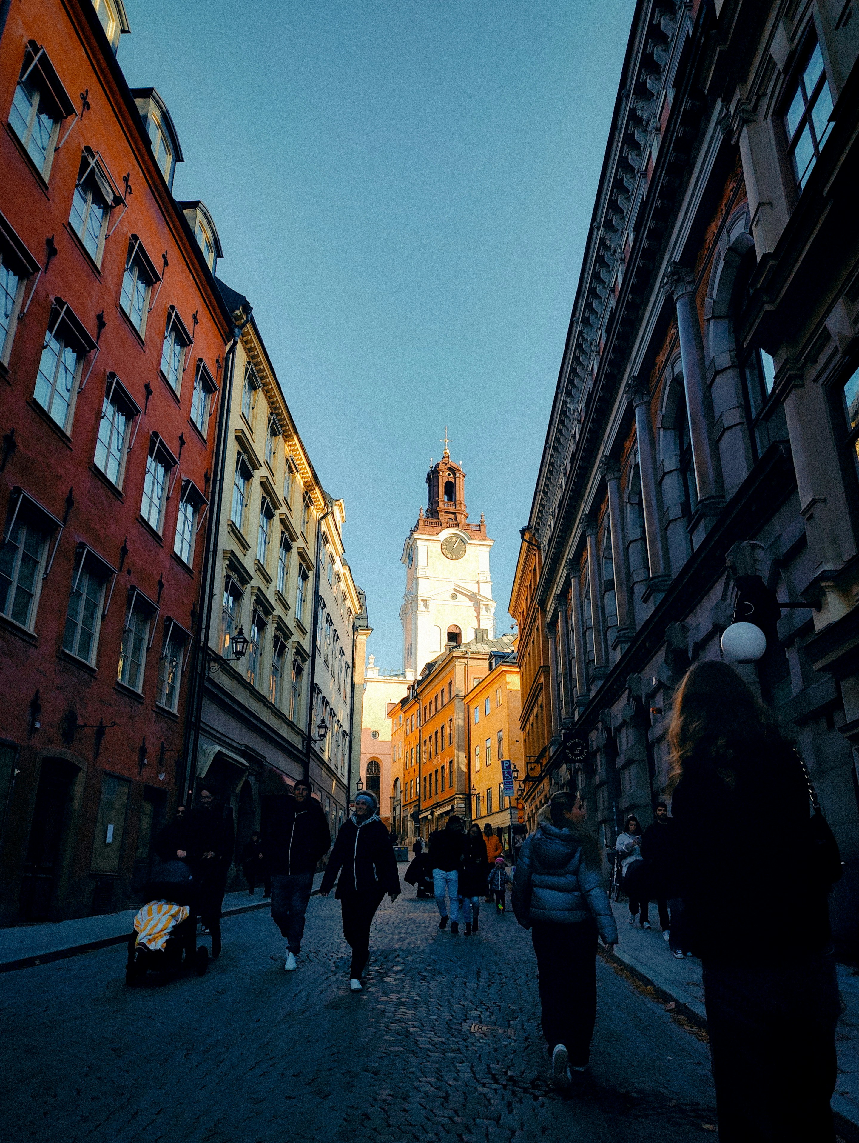 Charming narrow street lined with colorful buildings leading to a prominent clock tower. People leisurely walk along the cobblestone path.