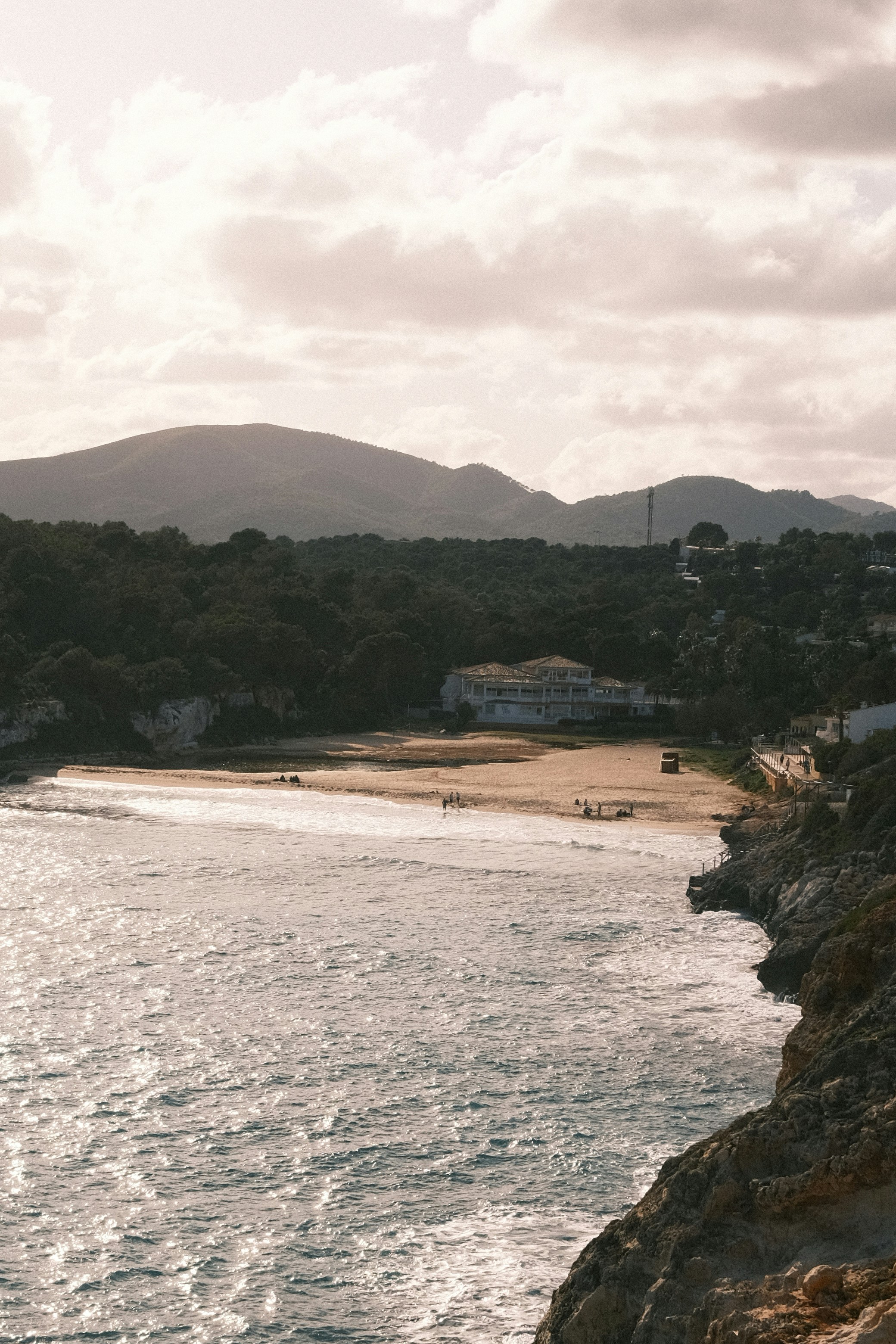 A tranquil beach scene with gentle waves lapping at the shore, surrounded by lush greenery and distant mountains under a cloudy sky.