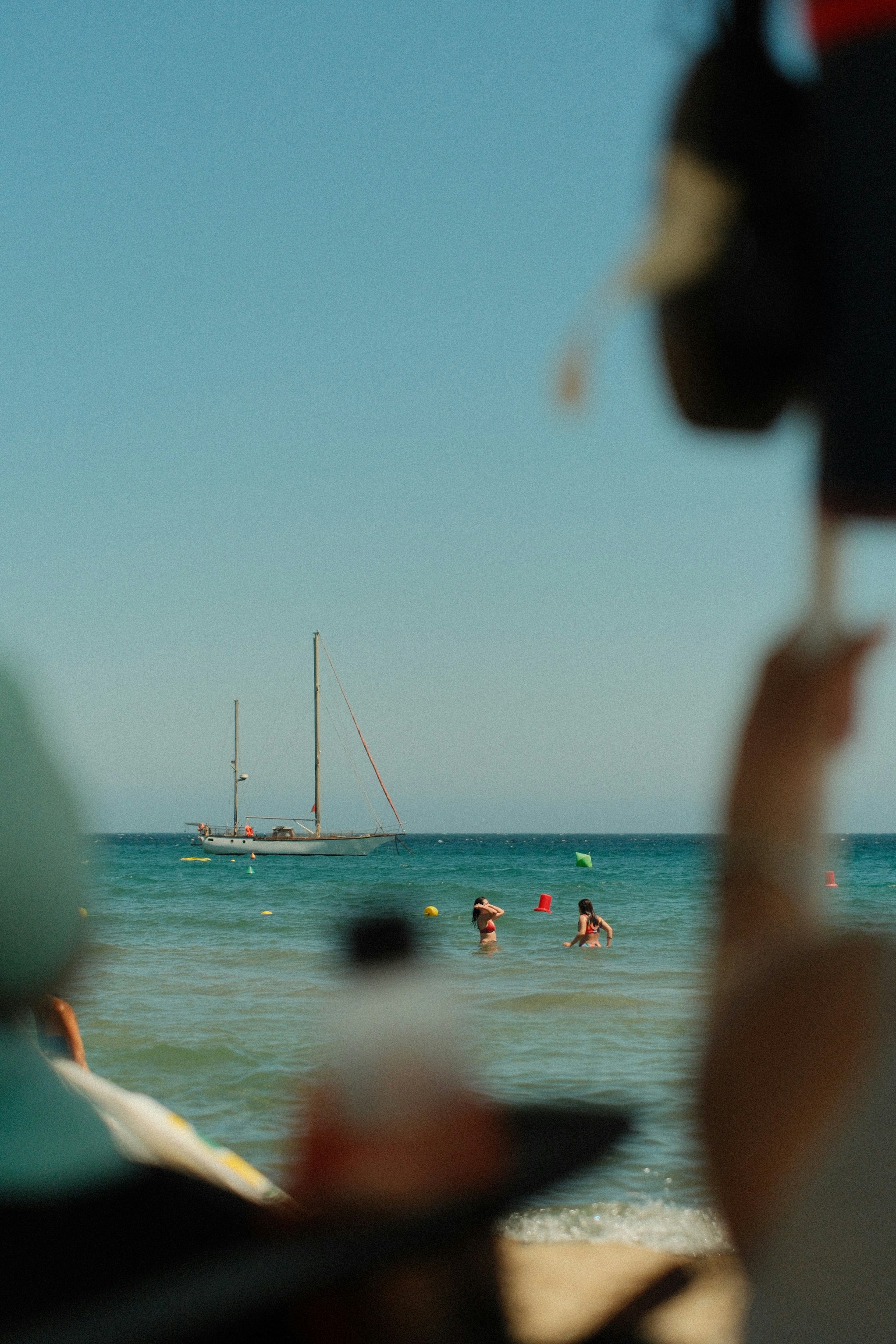 A classic summer scene captured with a warm, film-like quality. From a candid viewpoint on the shore, two women swim in the calm, turquoise sea while a sailboat is anchored in the distance under a clear blue sky. The blurry foreground frames the moment, giving it a personal, nostalgic feeling of a perfect holiday. | People swim near a sailboat on a sunny beach.