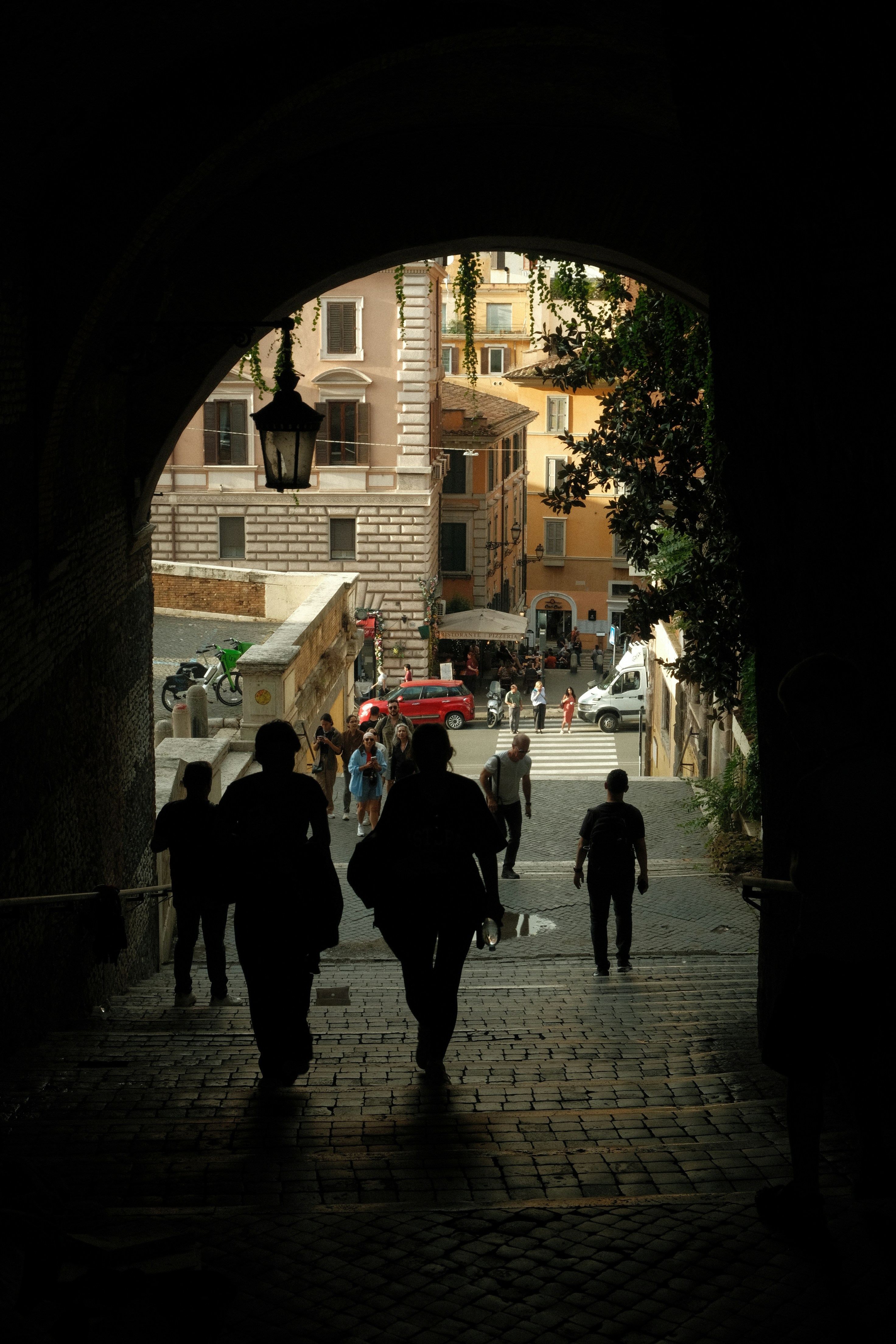 Silhouetted figures descending a stone staircase, framed by an archway, leading to a bustling street scene filled with pedestrians and vibrant architecture.