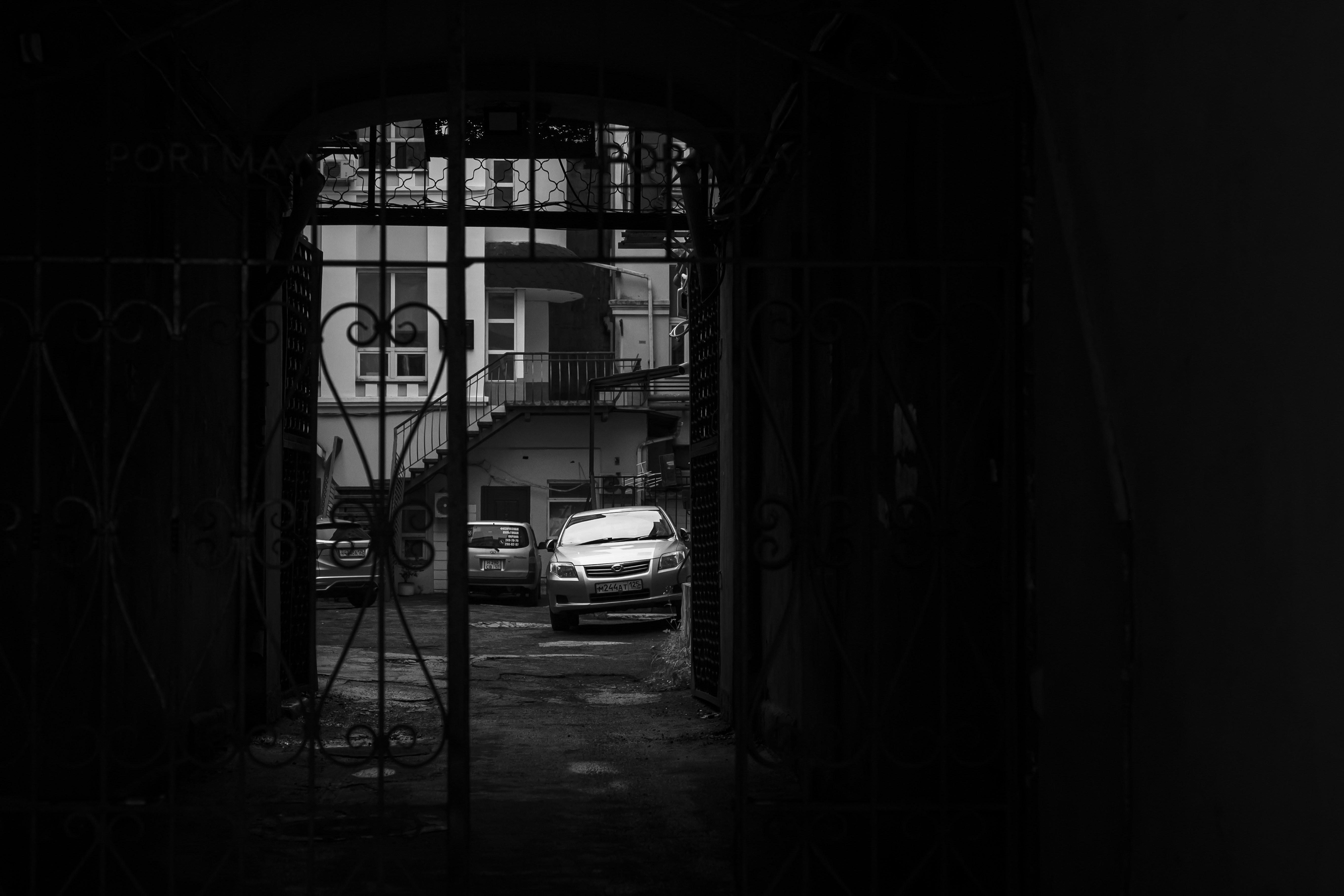 View through an archway with a lattice to the courtyard of a residential building with parked cars | A car and building are seen through a gate.