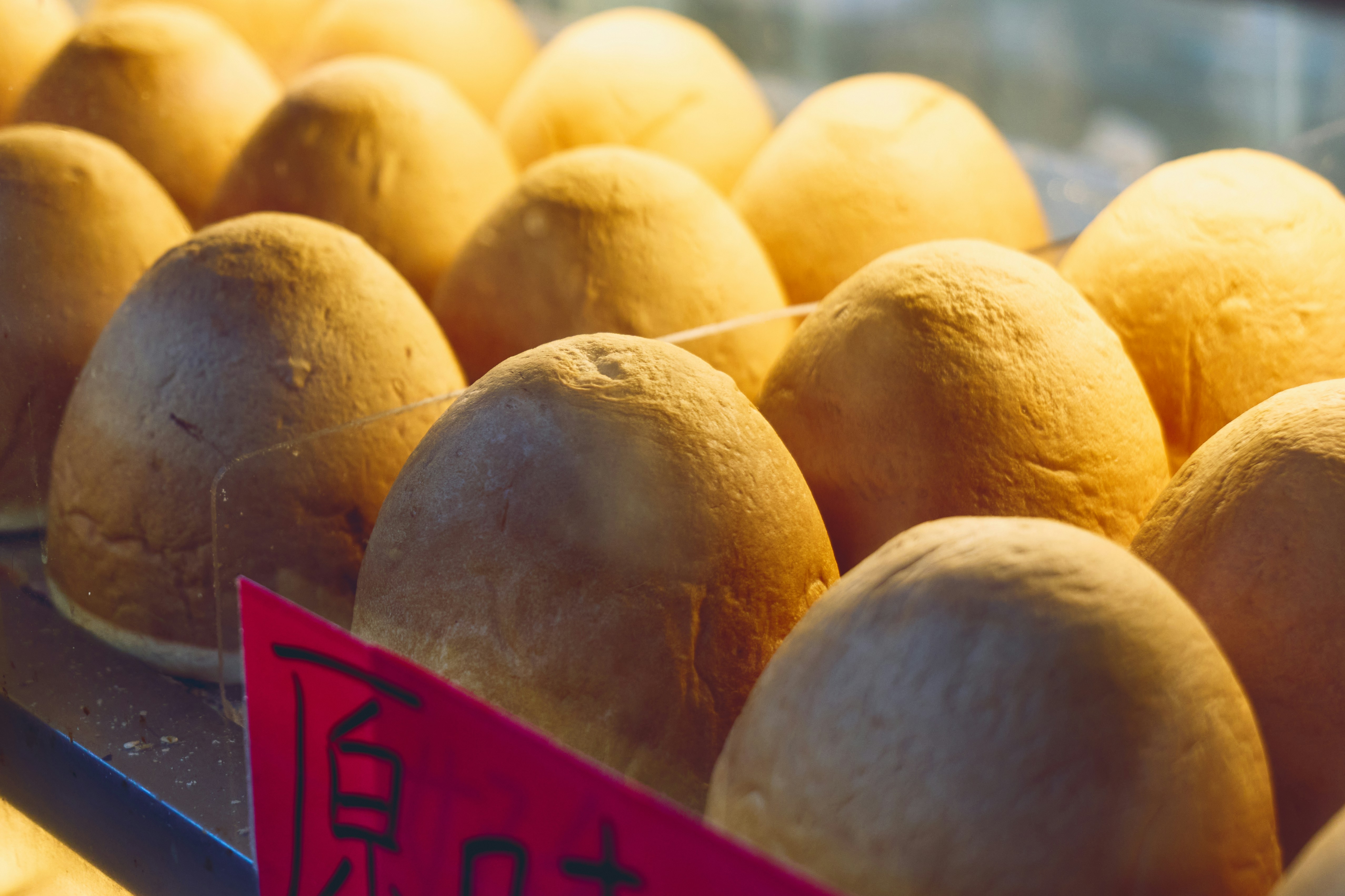 Freshly baked round bread rolls displayed in a bakery, showcasing their golden crusts and soft texture. A vibrant sign in the foreground adds to the scene.
