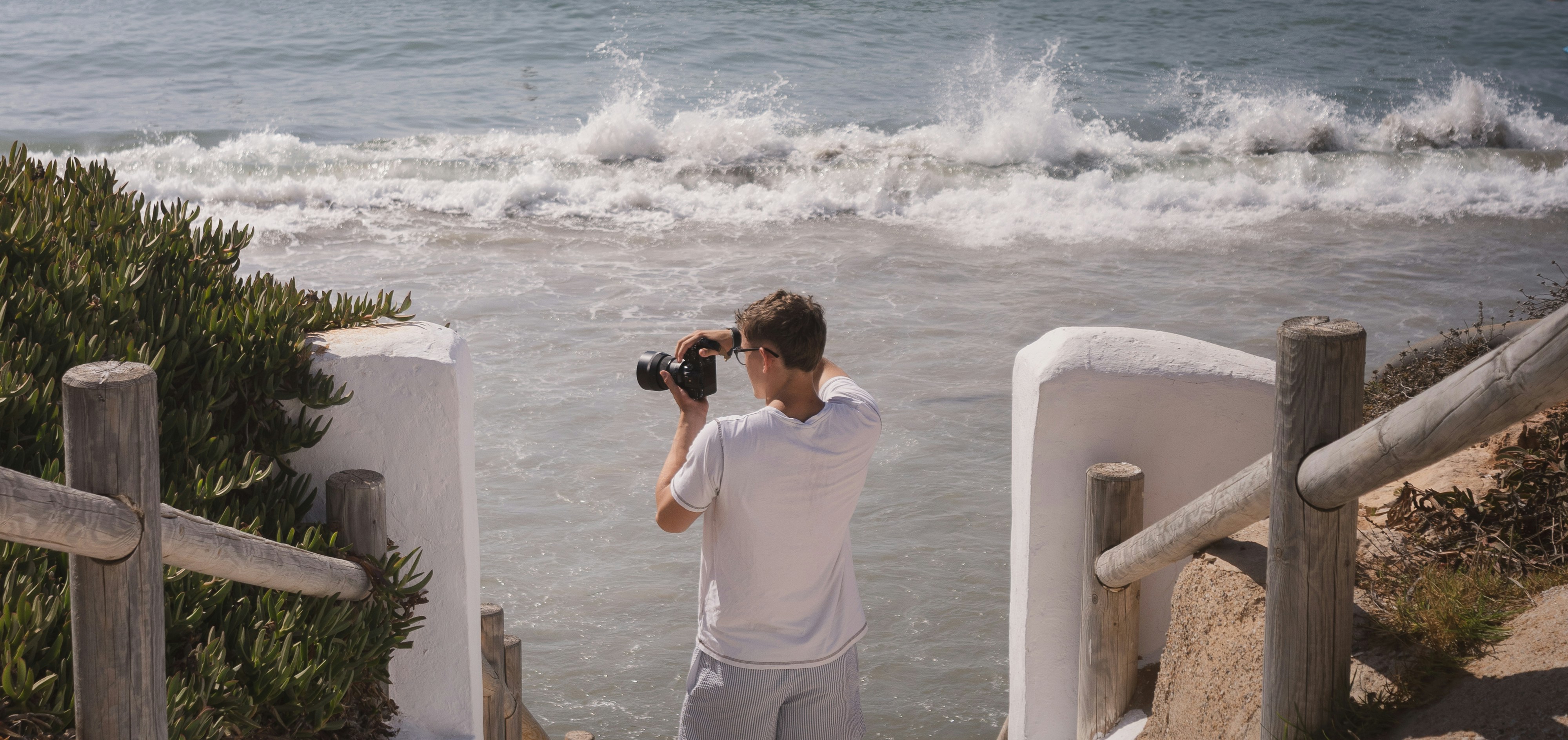 A man shooting with his camera while standing on the beach stairs. | A man films the ocean waves with a camera.