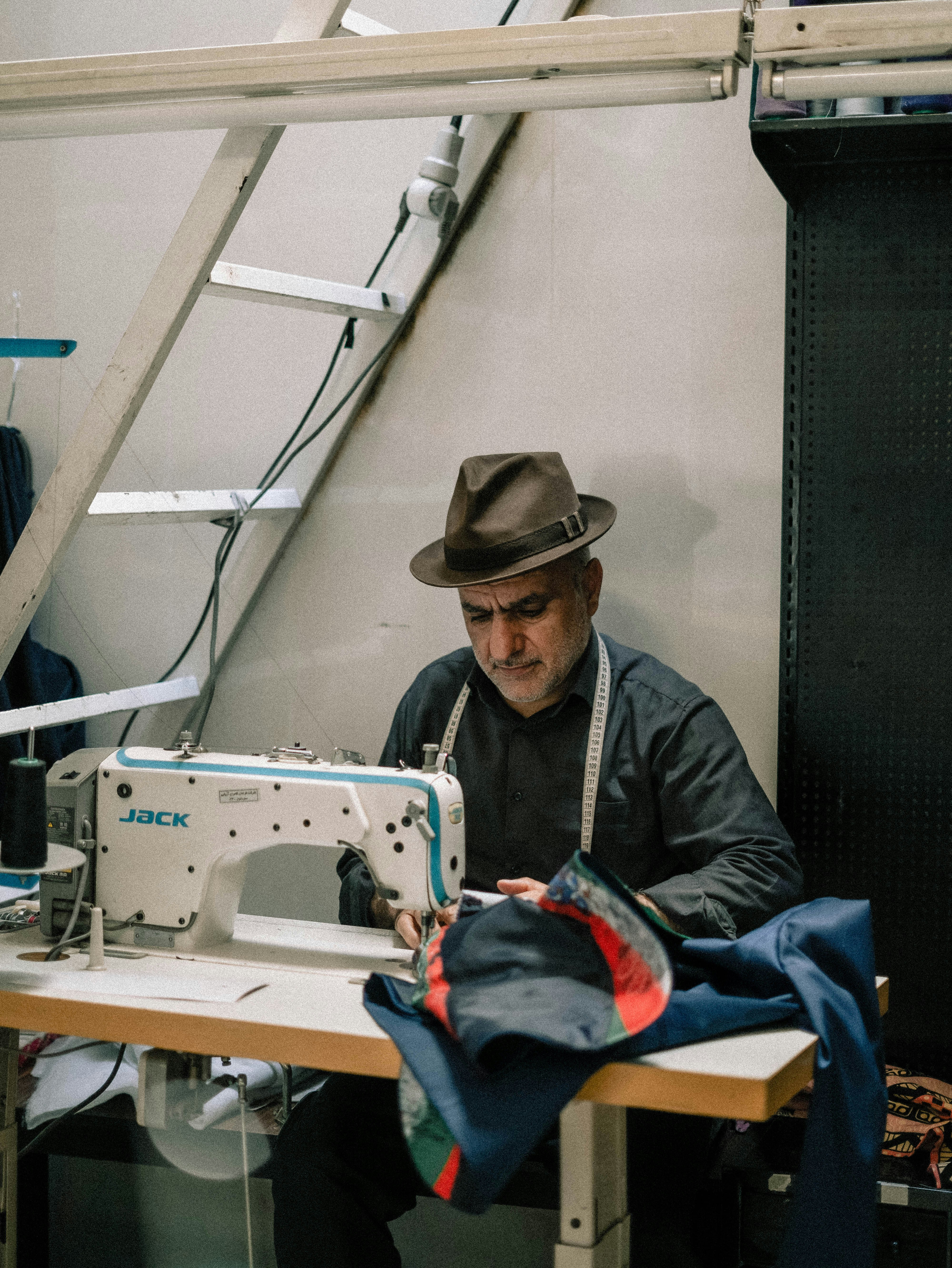 A skilled tailor focuses on his work at a sewing machine, surrounded by fabric and tools in a workshop setting.