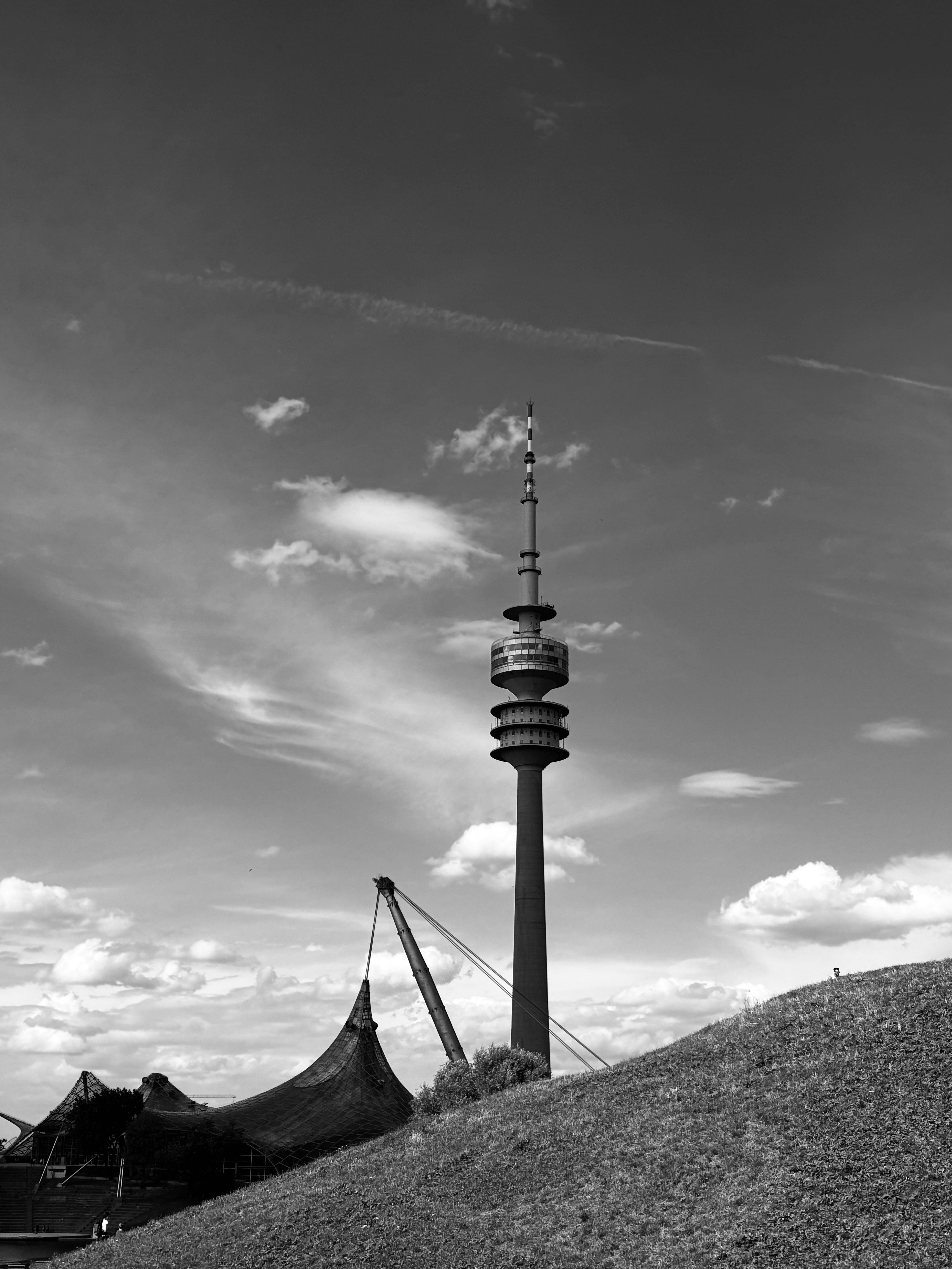 A towering broadcast antenna rises against a backdrop of dynamic clouds, with construction elements in the foreground. The scene's monochrome tones emphasize the contrast between nature and industry.