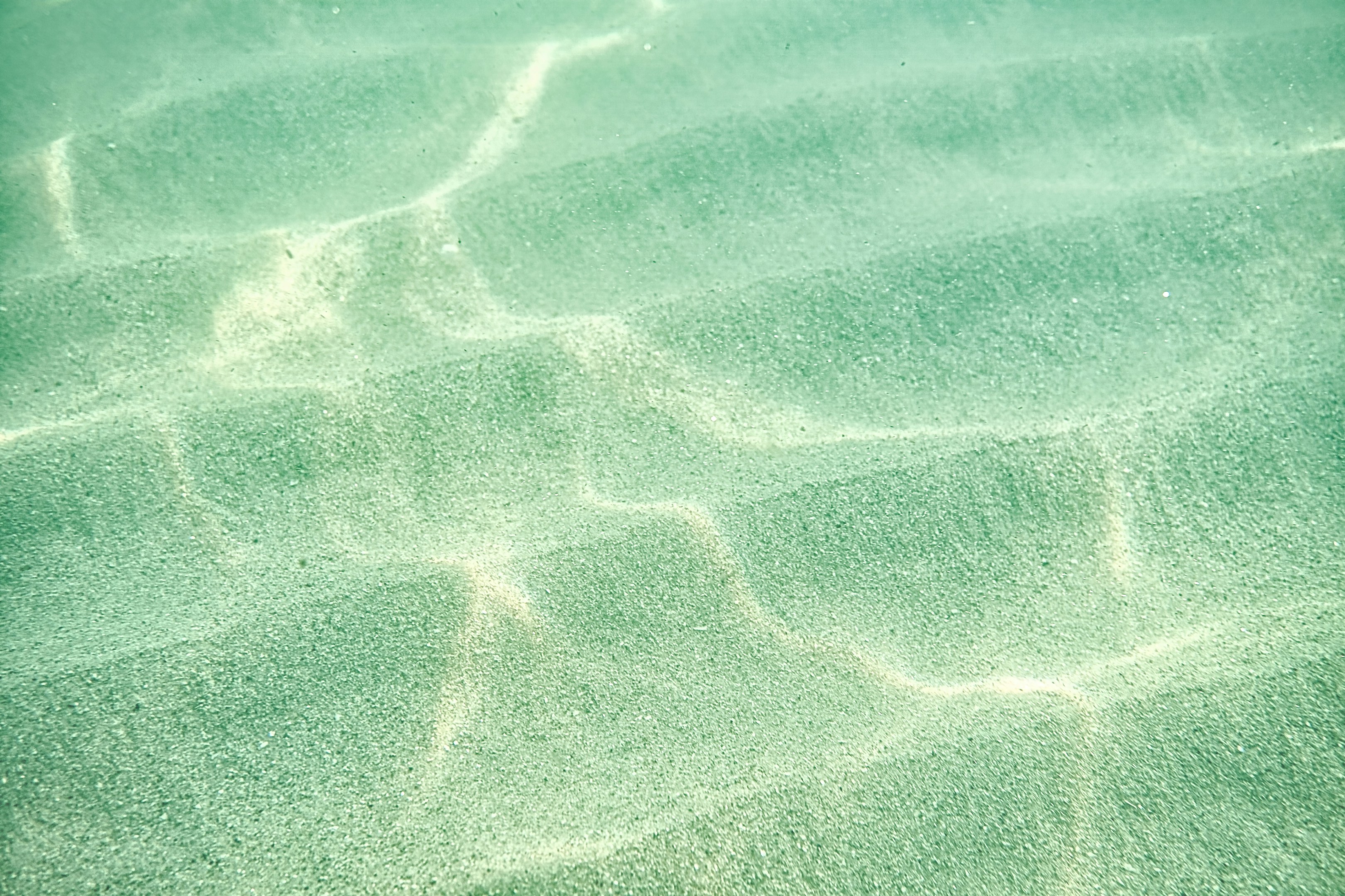 Gentle ripples and patterns on a sandy seabed illuminated by sunlight filtering through water.