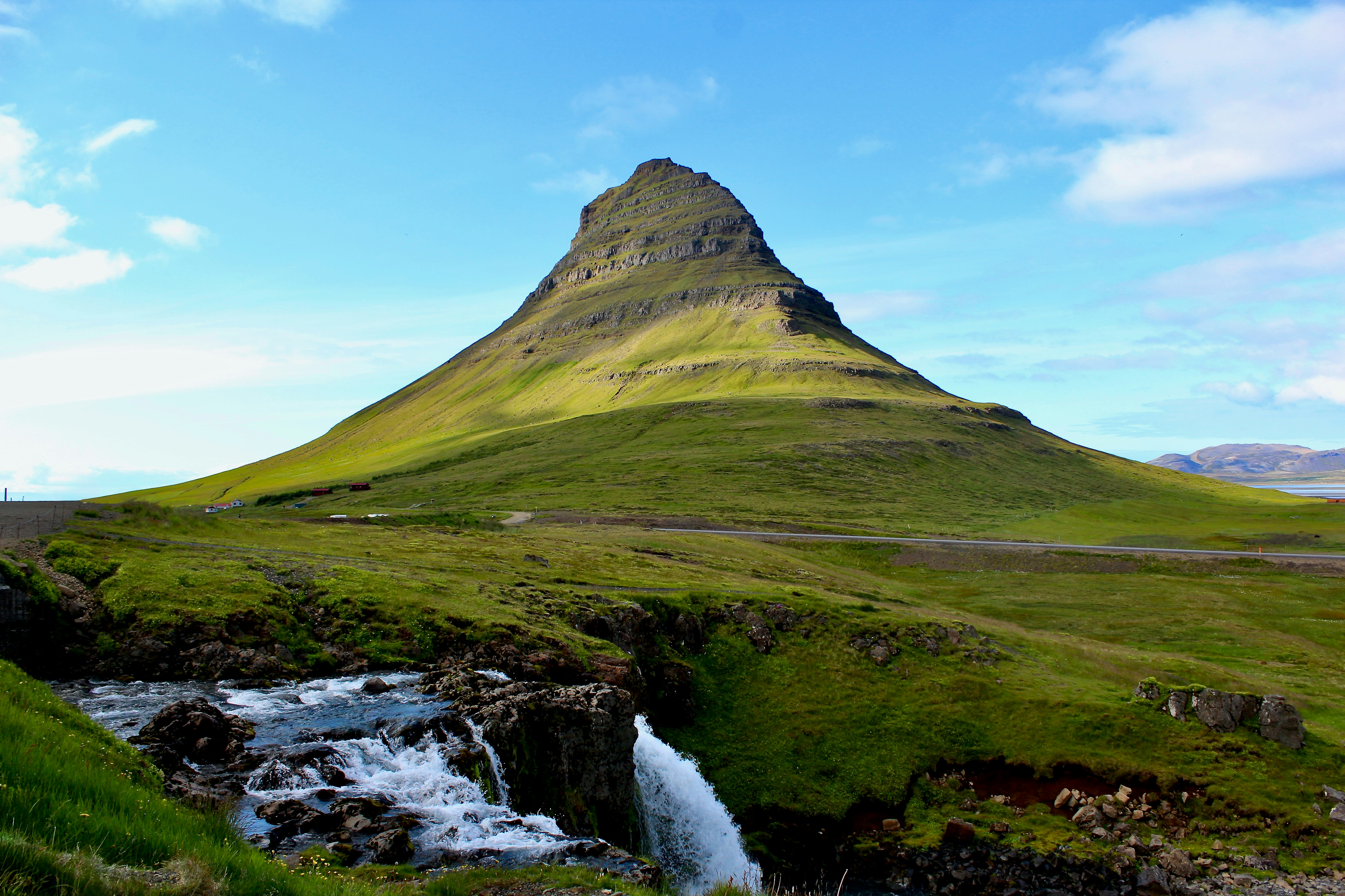 Kirkjufell mountain towers over a cascading waterfall, framed by lush greenery and a clear blue sky.