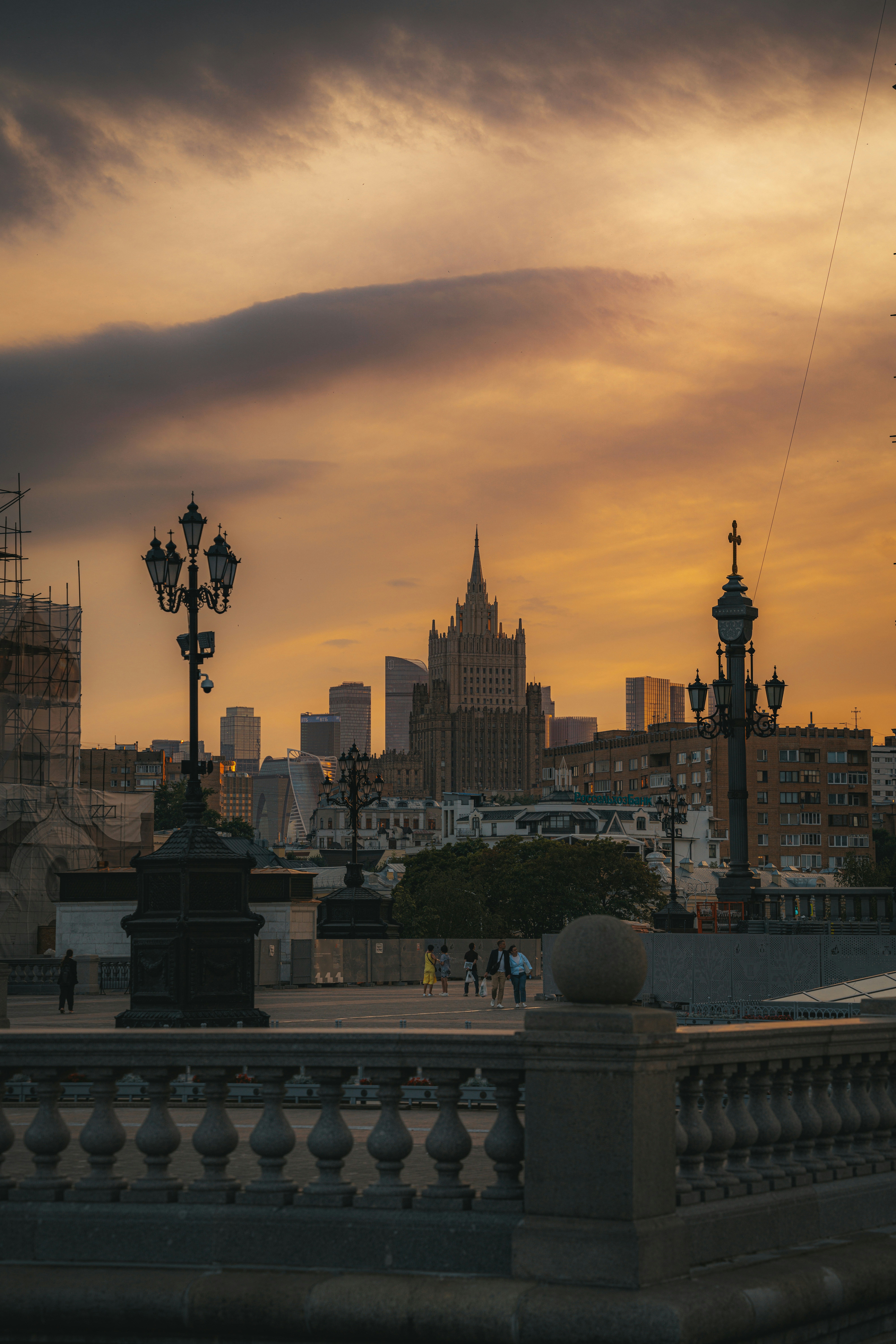 The skyline features a prominent tower amidst a backdrop of dramatic clouds at sunset, with street lamps and pedestrians in the foreground.