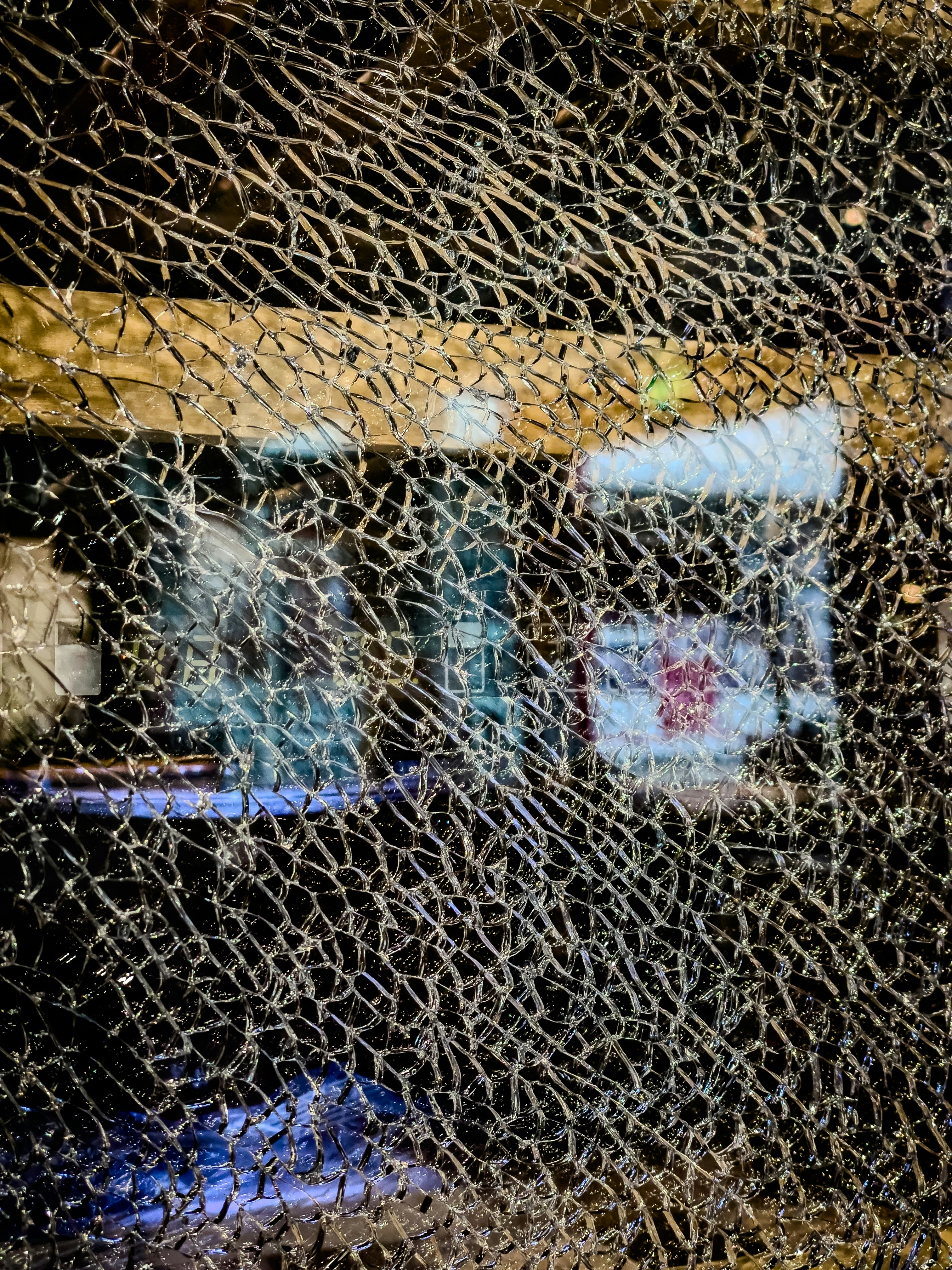 A close-up view of a shattered safety glass panel shows intricate crack patterns spreading across the surface. Reflections of an indoor space, including wooden beams and light sources, are visible behind the fractured glass. | Shattered glass reveals a blurred storefront.