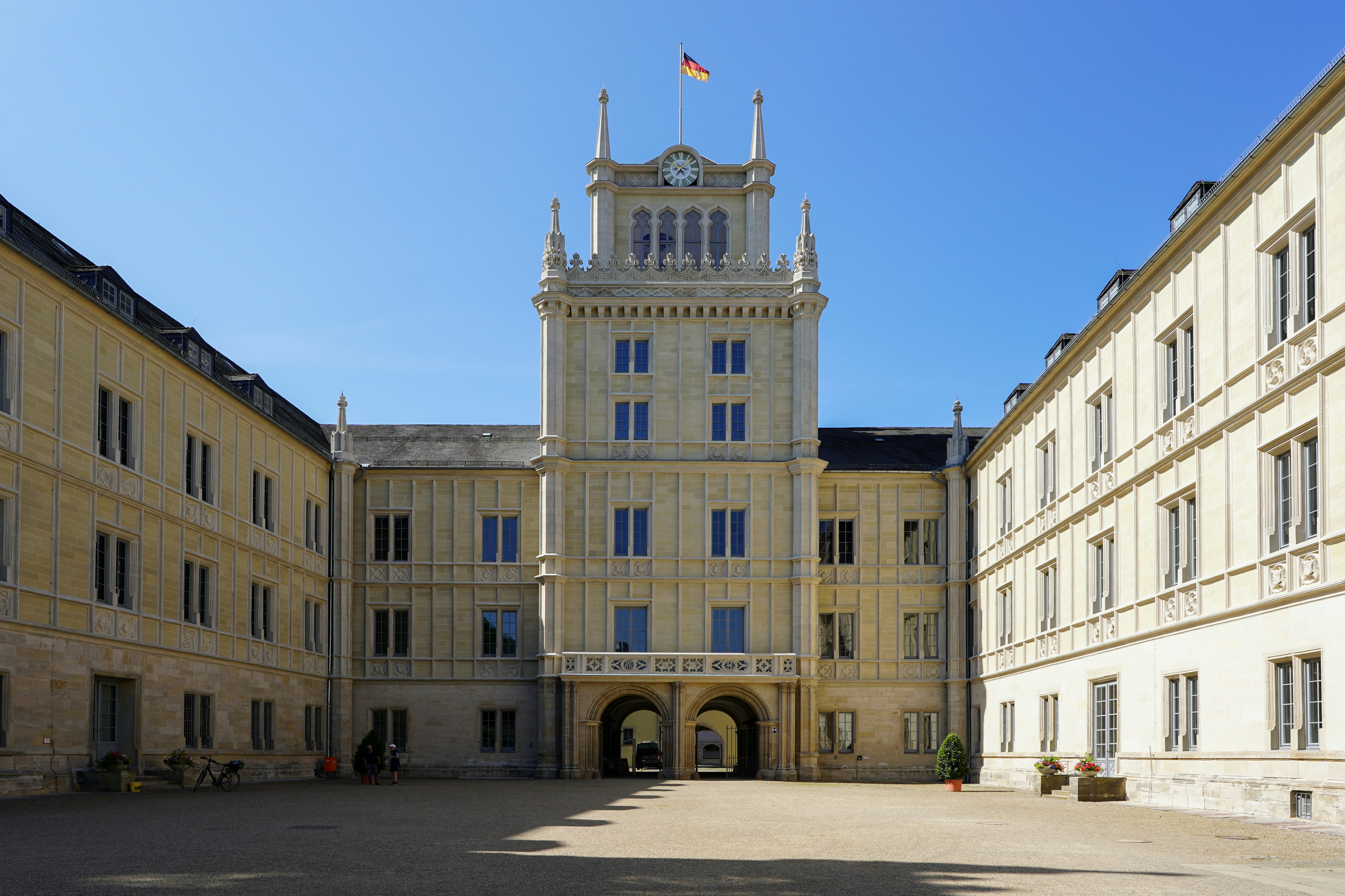 A grand building with a clock tower stands prominently in a courtyard, flanked by symmetrical wings of stone. The German flag waves atop, adding a touch of national pride.