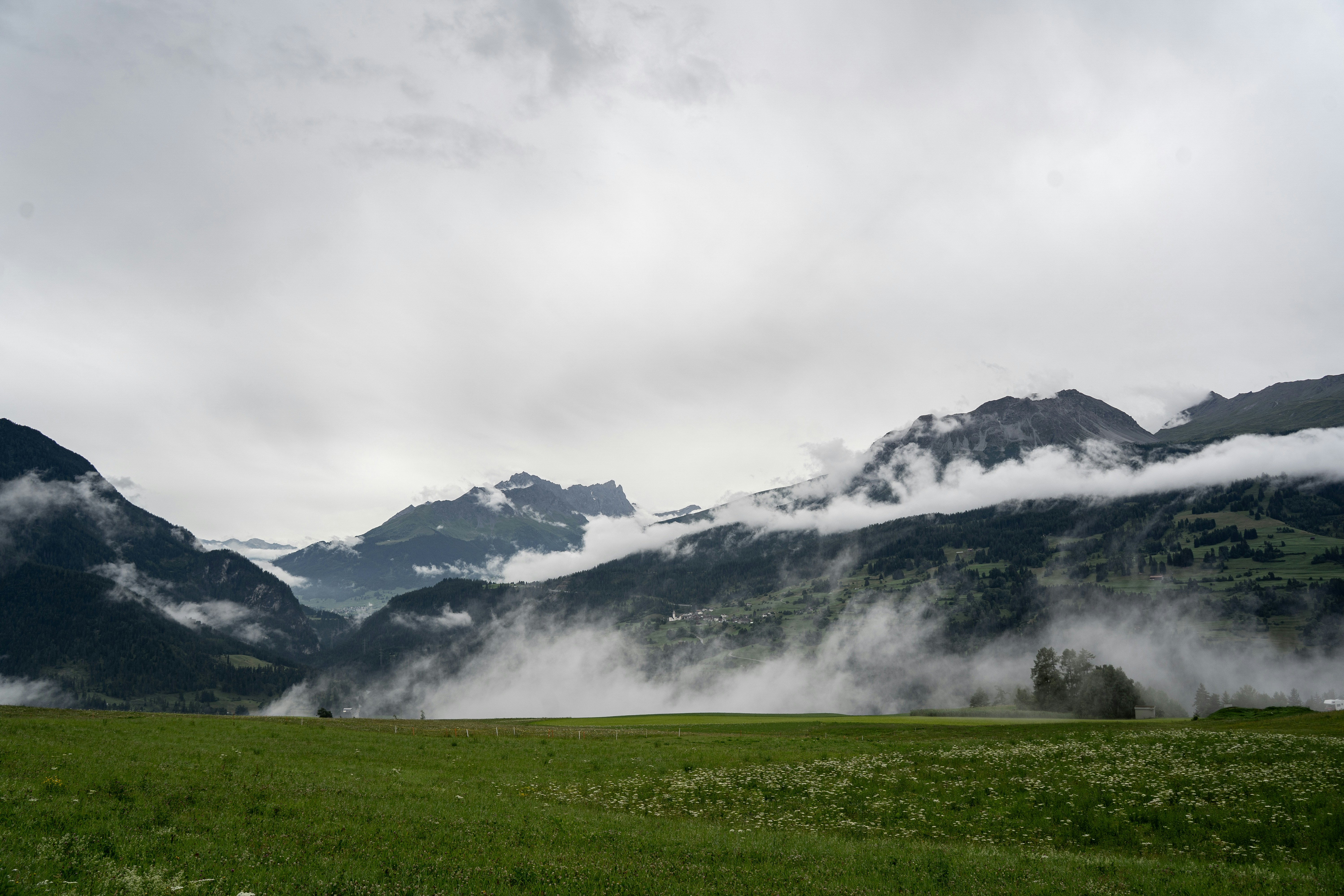 Low-hanging clouds drift through a moody alpine valley in Switzerland, with mist weaving between dark forested slopes and green meadows under a dramatic overcast sky. The scene captures a calm, ethereal beauty that blurs the line between earth and sky. | Mountains are shrouded in mist and clouds.