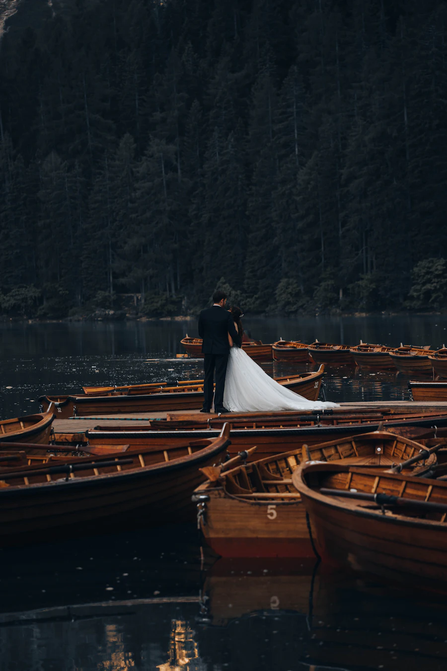 A bride in a flowing white dress and a groom in a dark suit stand together on a dock surrounded by wooden rowboats, with a dark forested shoreline behind them.