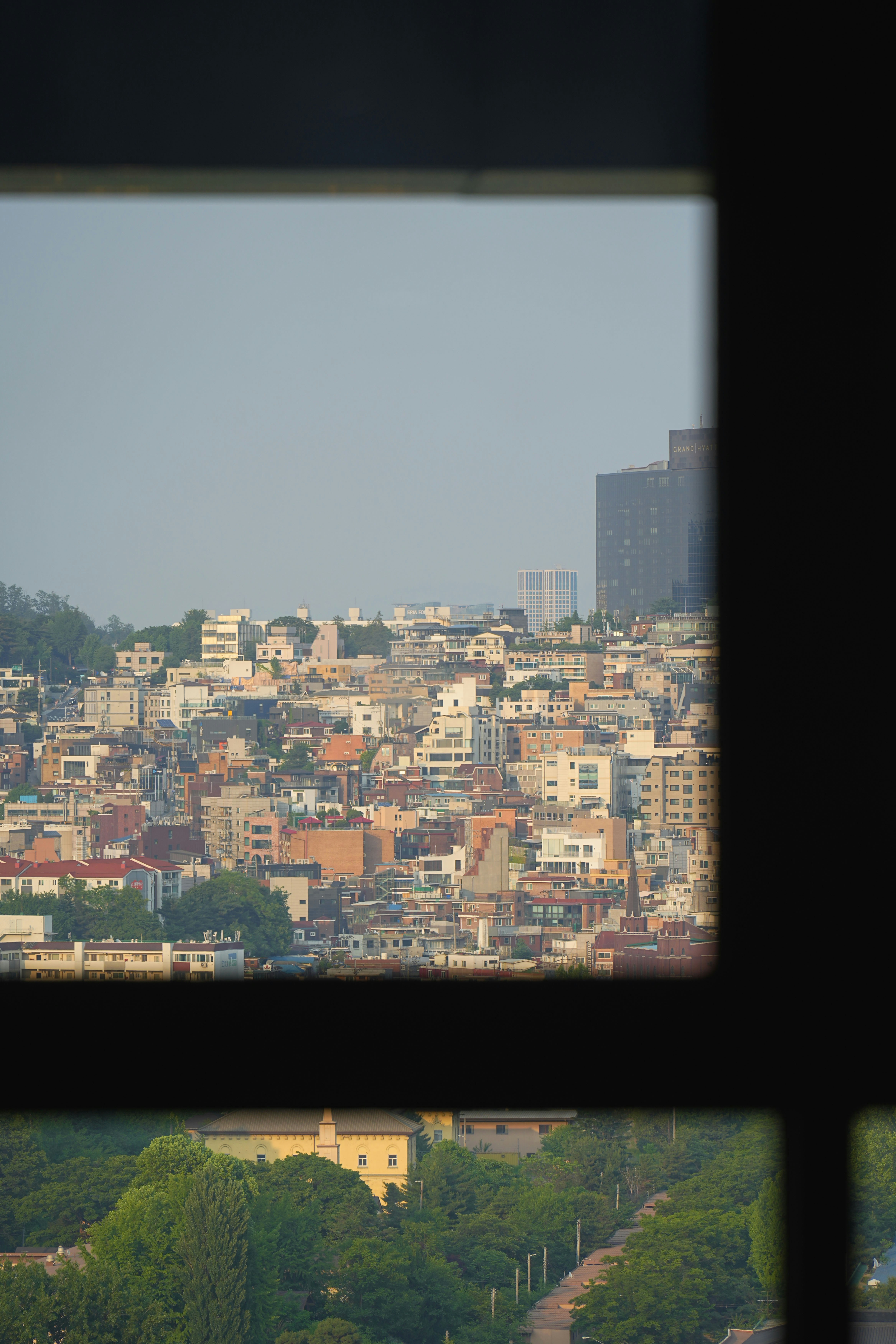 A panoramic view of a cityscape framed by a window, showcasing a blend of modern and traditional architecture amid lush greenery.