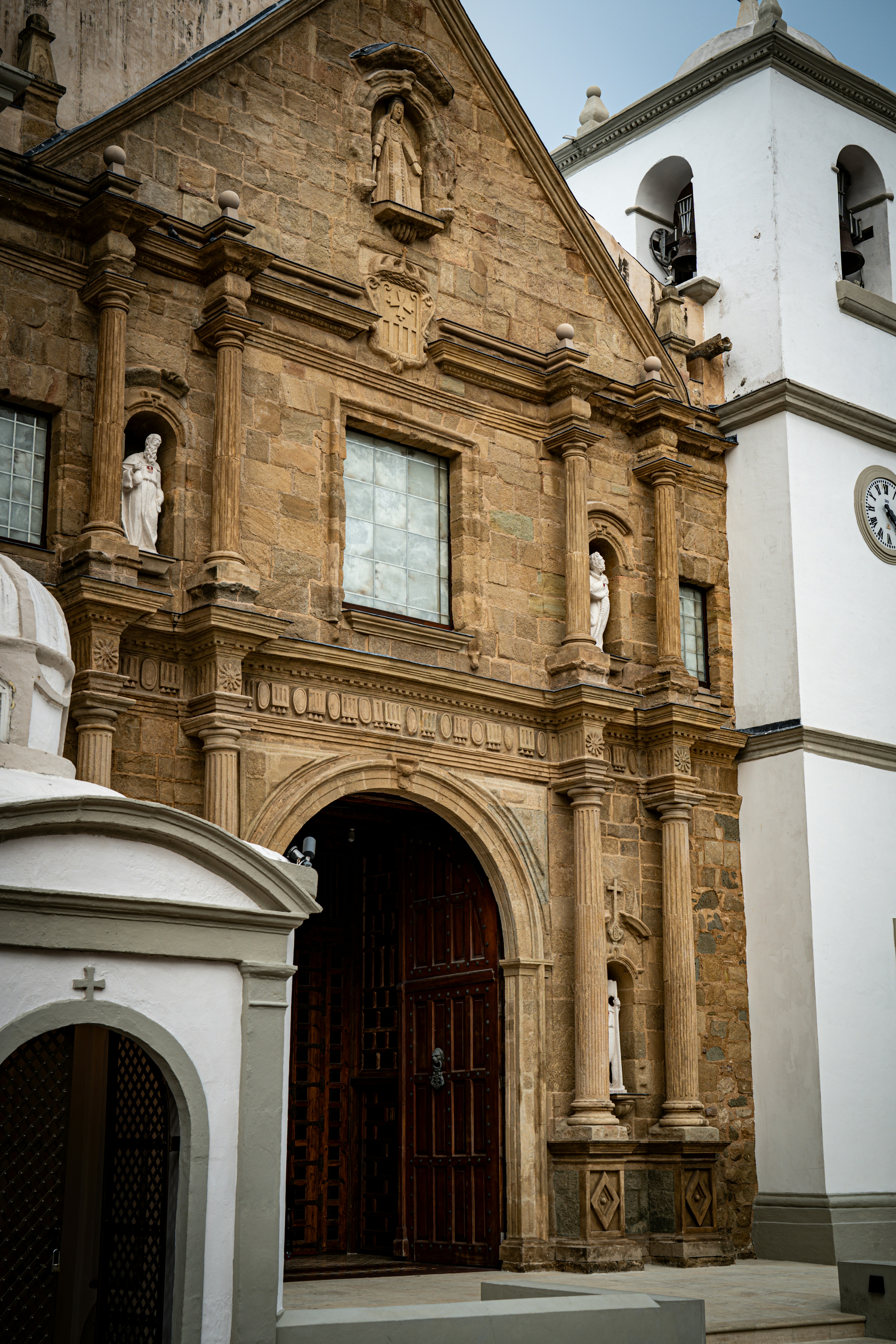 Intricate stone facade of a historic church featuring statues and detailed carvings, complemented by a neighboring structure with a clock tower.
