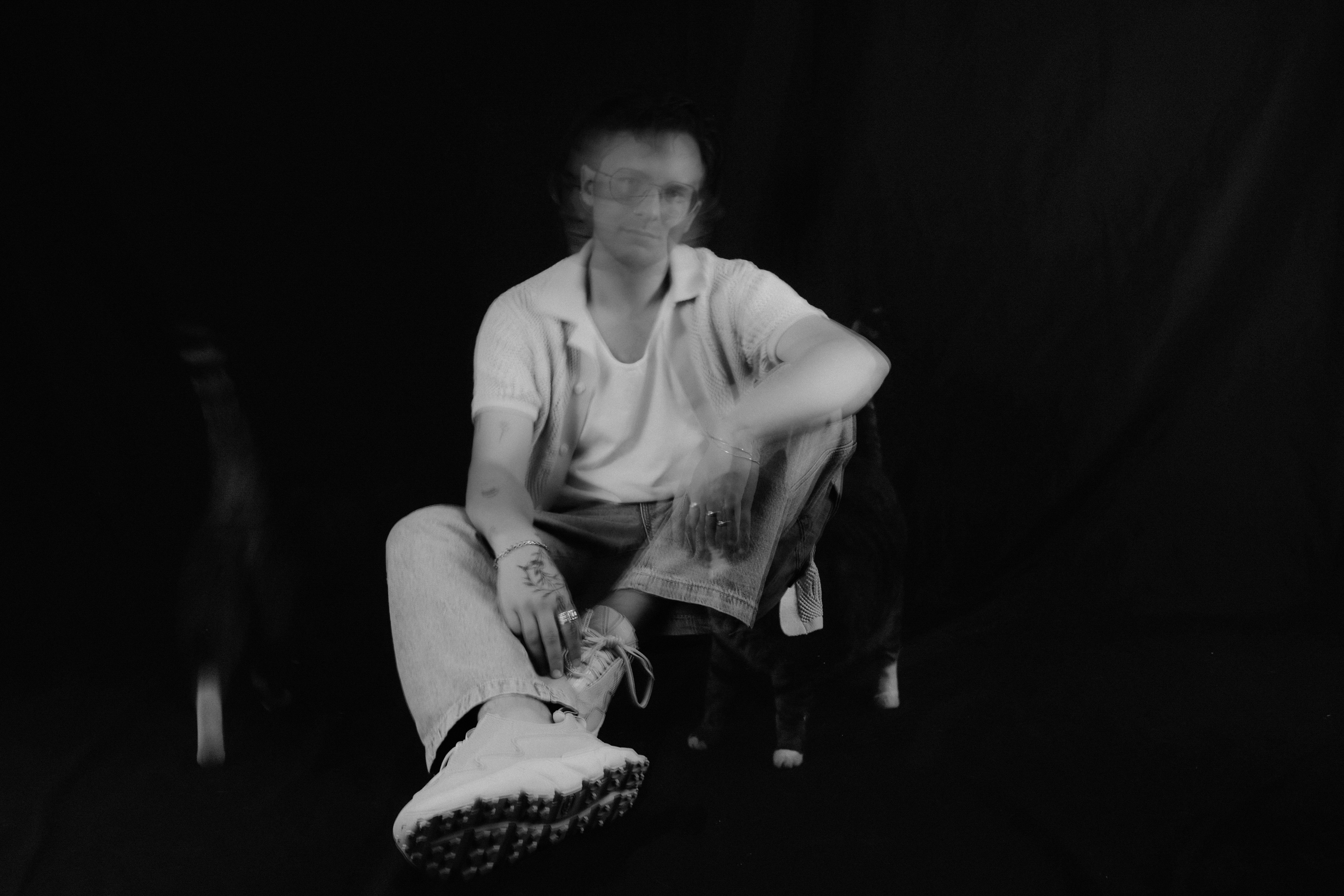 A person seated on the floor, partially obscured by motion blur, with two cats nearby against a dark backdrop.
