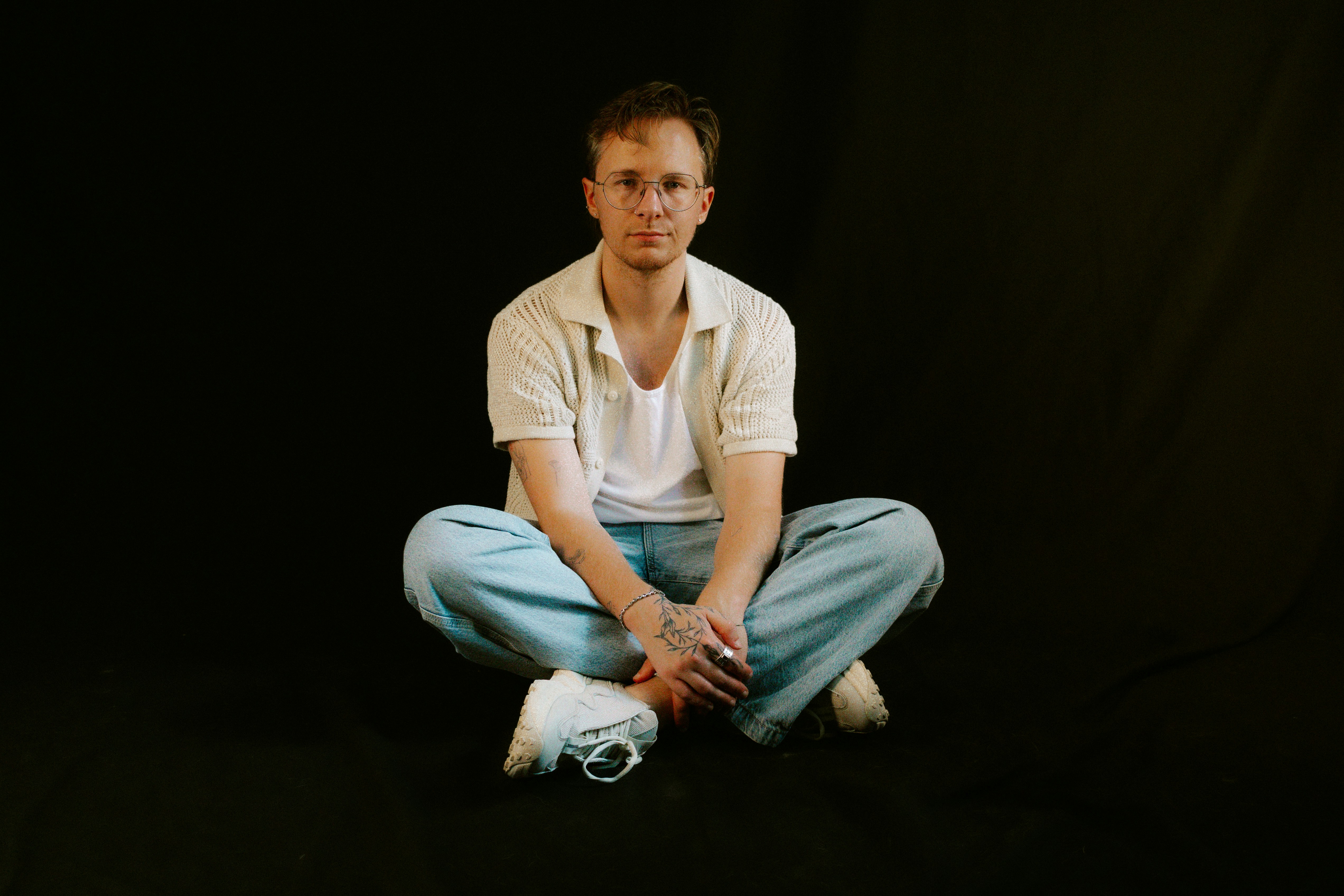 A person seated cross-legged on a dark background, wearing casual attire and glasses, exuding a sense of calm reflection.