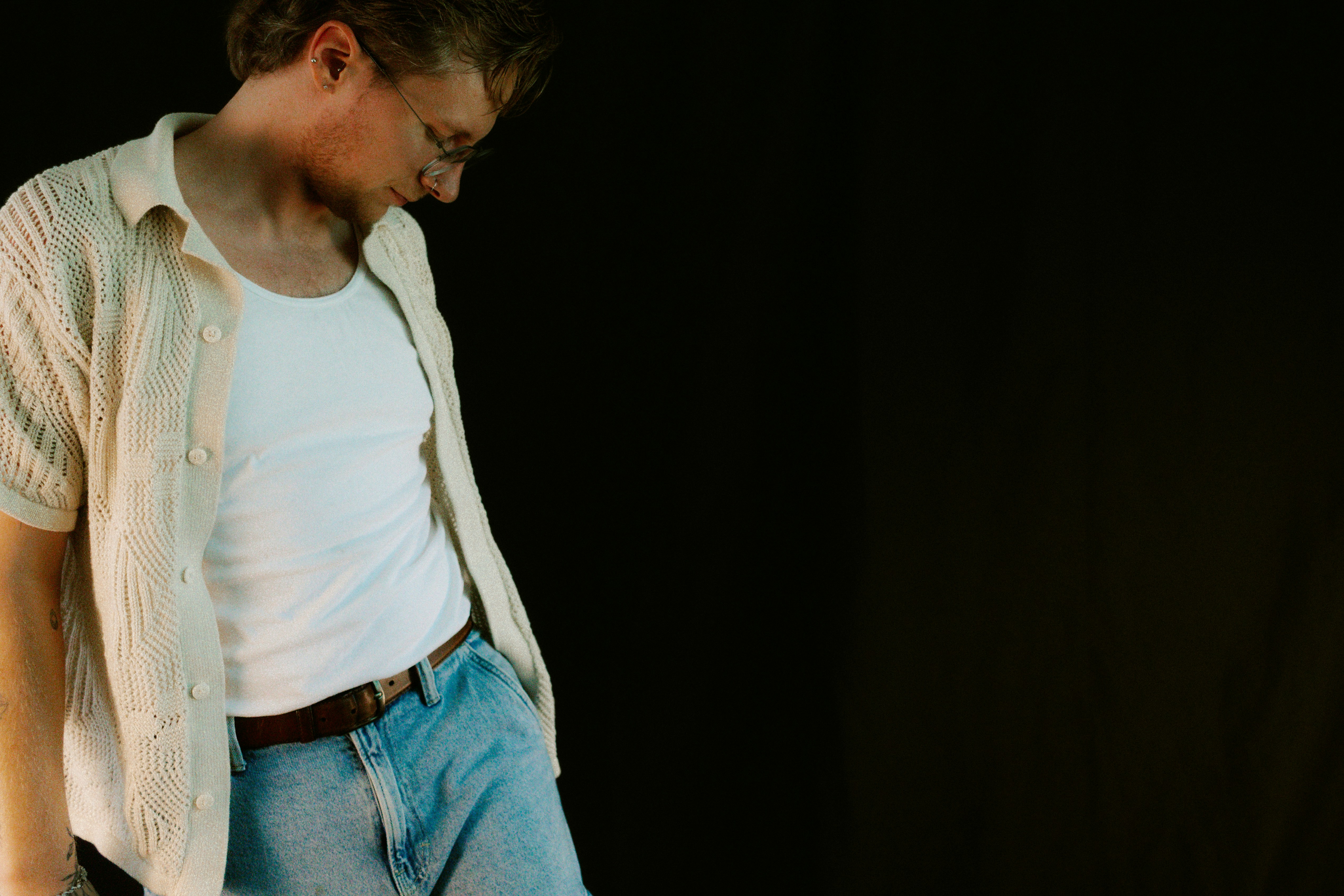 A man dressed in a light cardigan and tank top gazes downward against a dark backdrop, embodying a moment of introspection.
