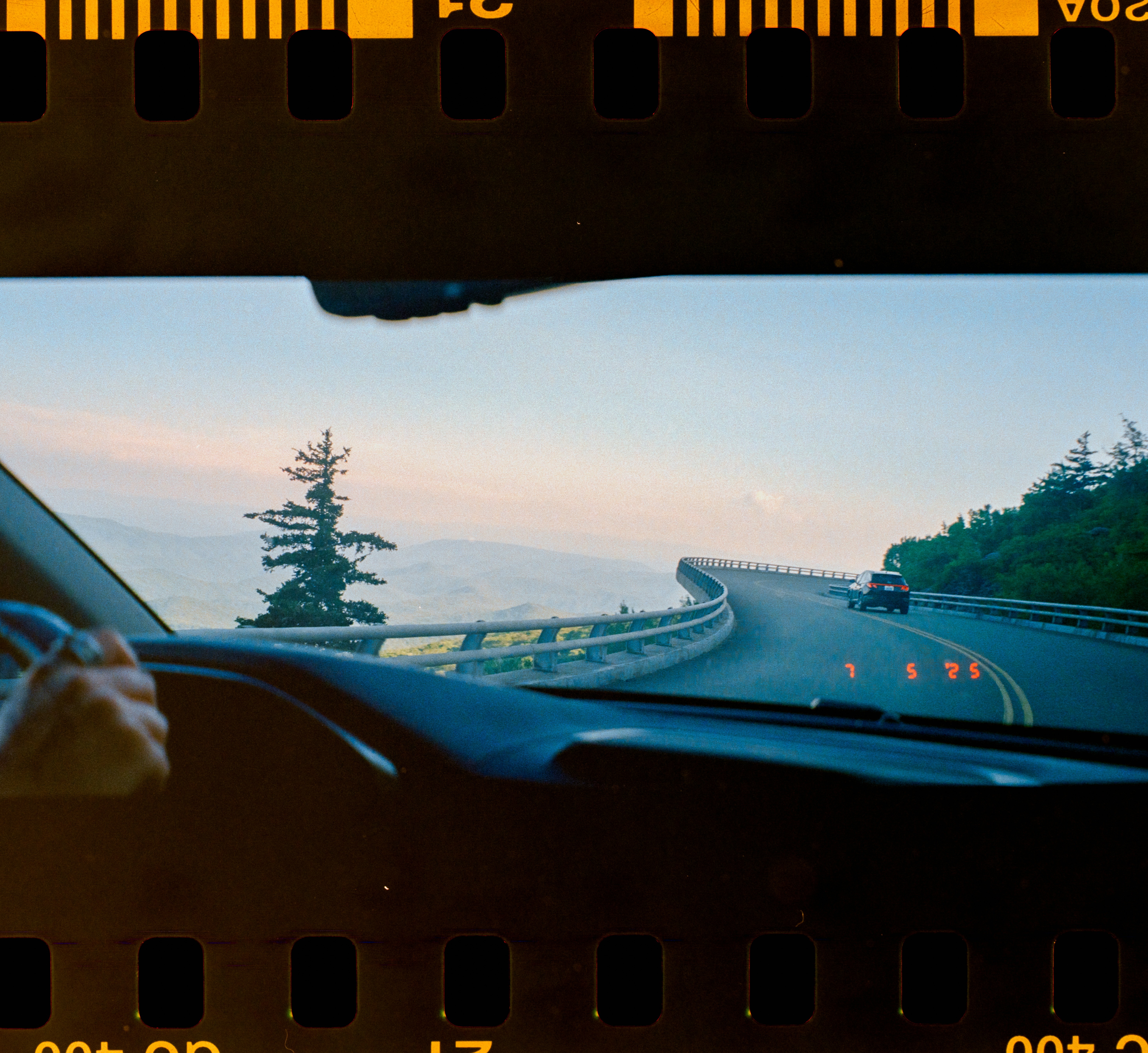 View from inside a car capturing a winding road alongside lush trees and distant mountains during twilight.