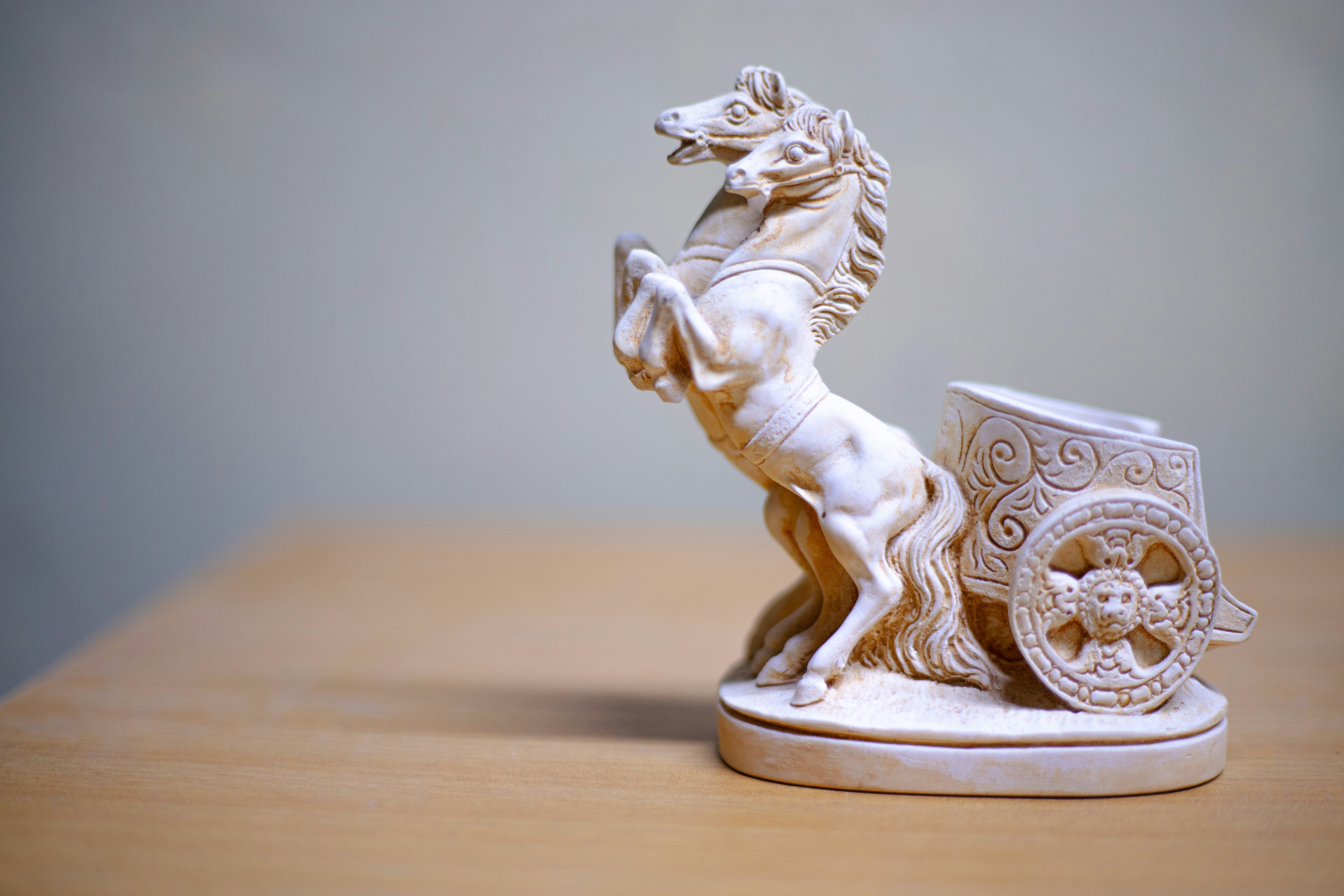 A detailed white sculpture of a Roman-style horse rearing up while pulling an ornate chariot. Crafted with precision, the muscular form of the horse and the intricate wheels of the carriage evoke classical elegance. Set on a light wooden surface against a soft gray background, the scene blends historical symbolism with minimalist presentation. | A decorative statue of a horse and chariot.