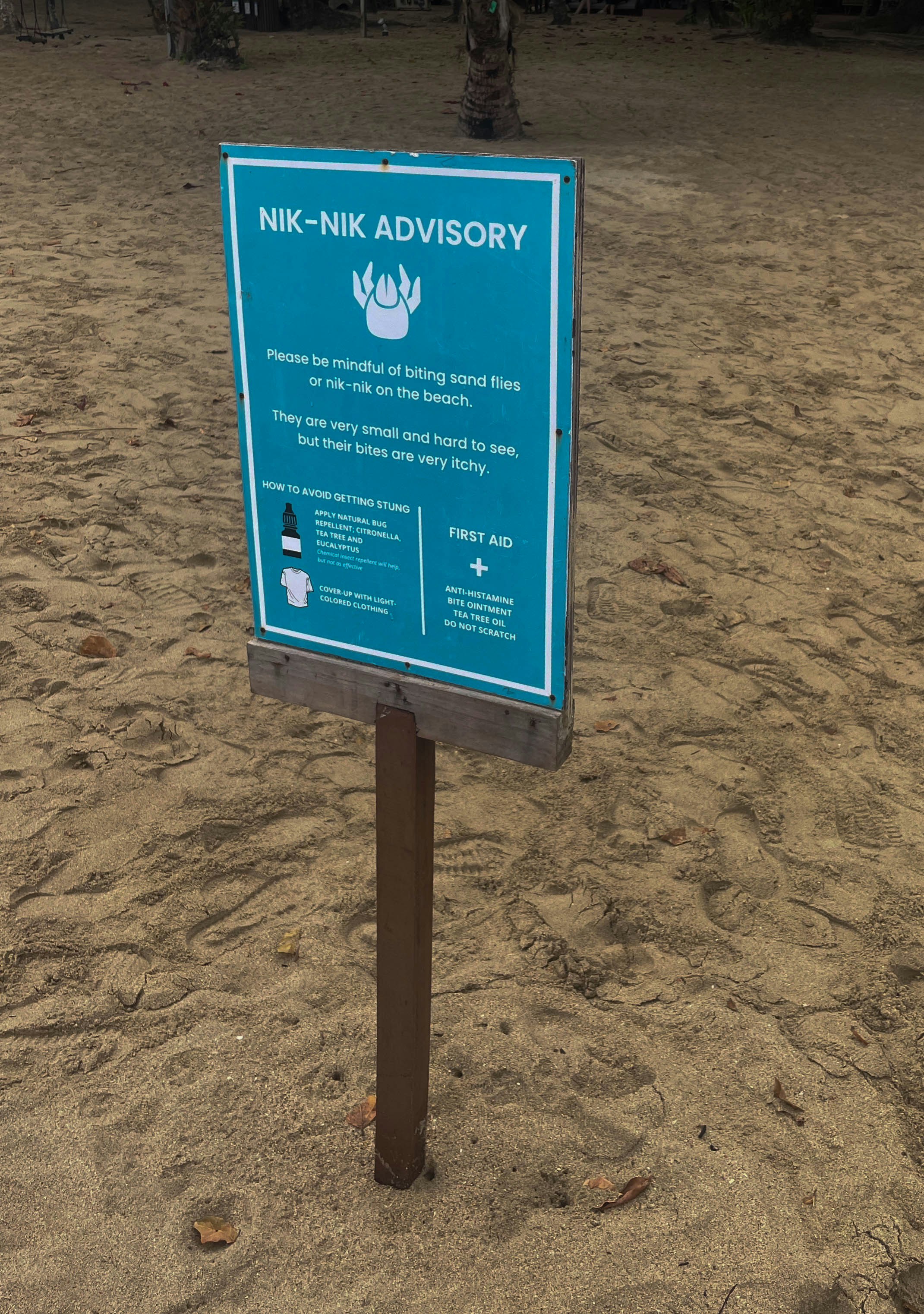 An informational sign posted on a sandy beach advises visitors about the presence of biting sand flies, known locally as nik-nik. It includes prevention tips and first aid recommendations for dealing with bites. | Sign warns of "nik-nik" bites on the beach.