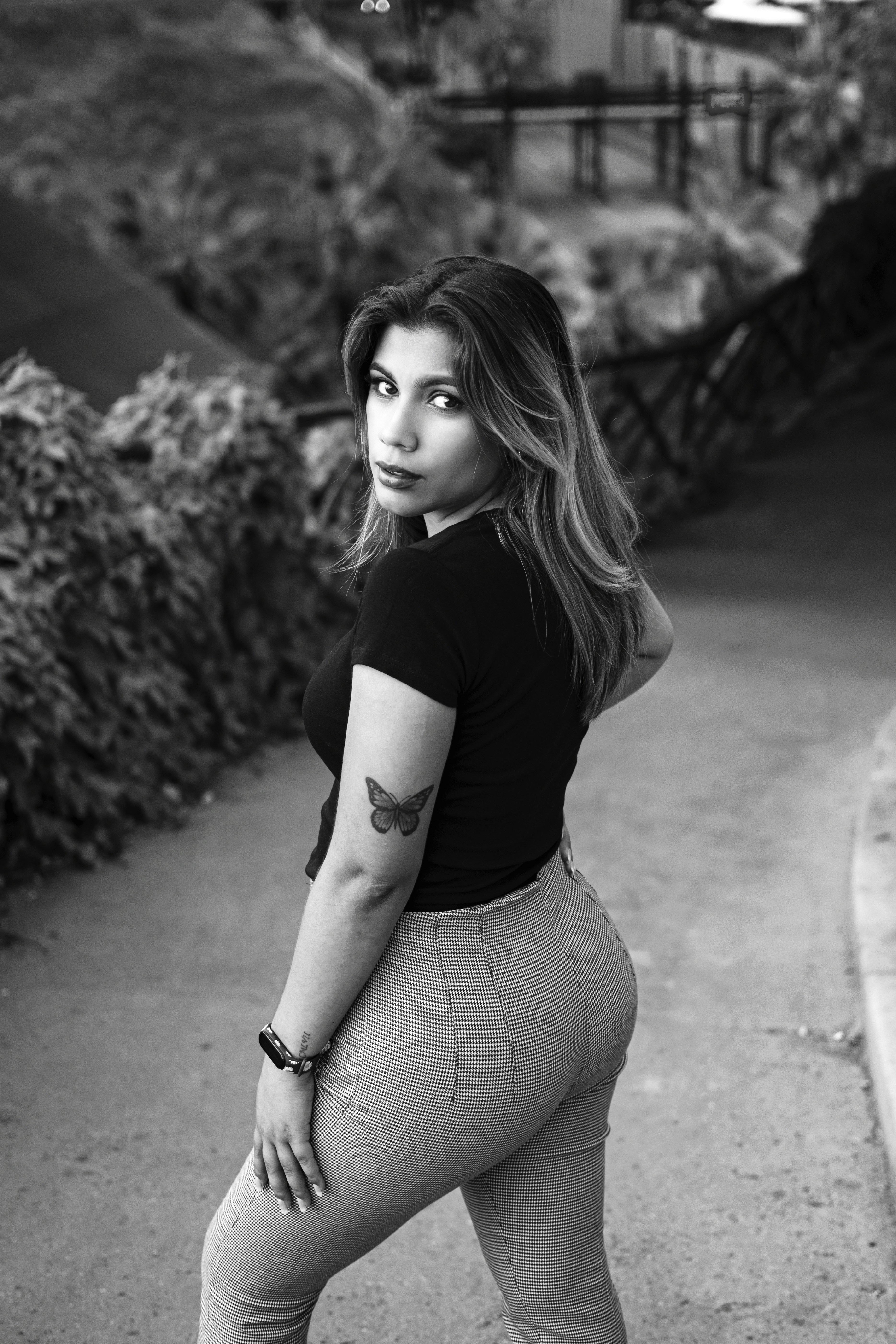 A young woman glances back while walking along a path surrounded by lush greenery, showcasing her butterfly tattoo. Black and white tones emphasize the mood of the scene.