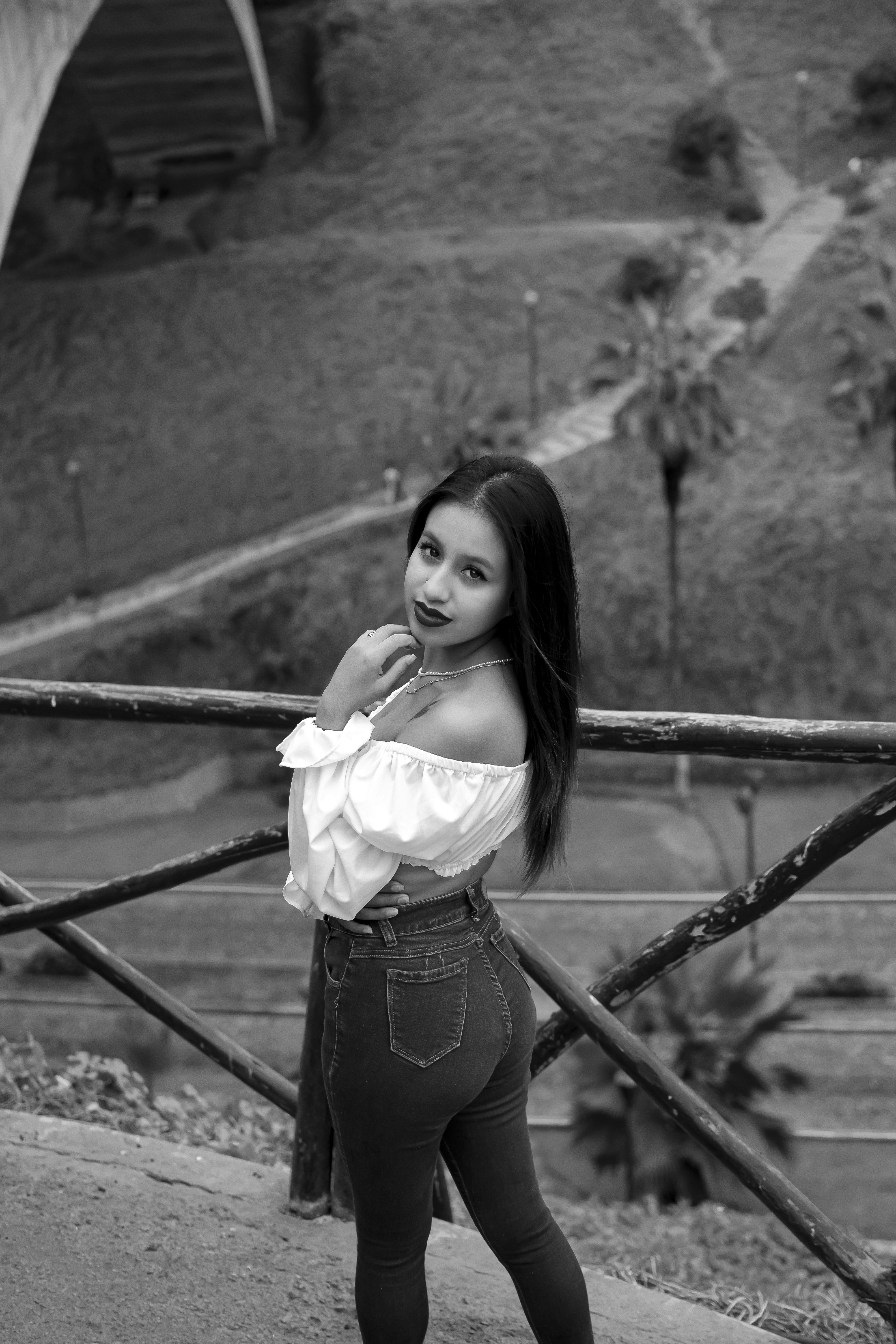 A young woman stands gracefully by a railing, her confident pose complemented by flowing hair and a stylish outfit, set against a blurred natural backdrop.