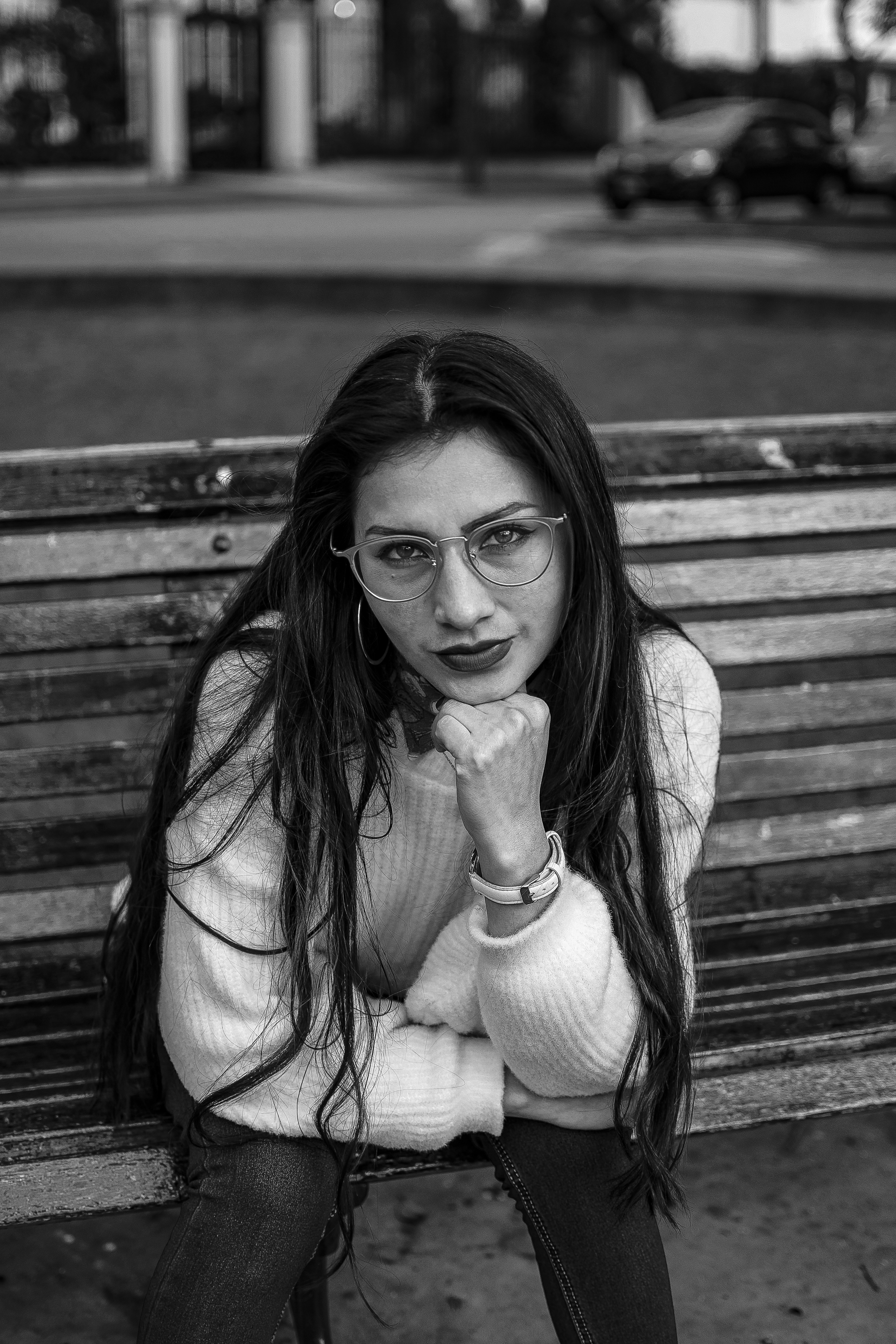 A young woman with long hair and glasses rests her chin on her fist while seated on a park bench, exuding a thoughtful demeanor.